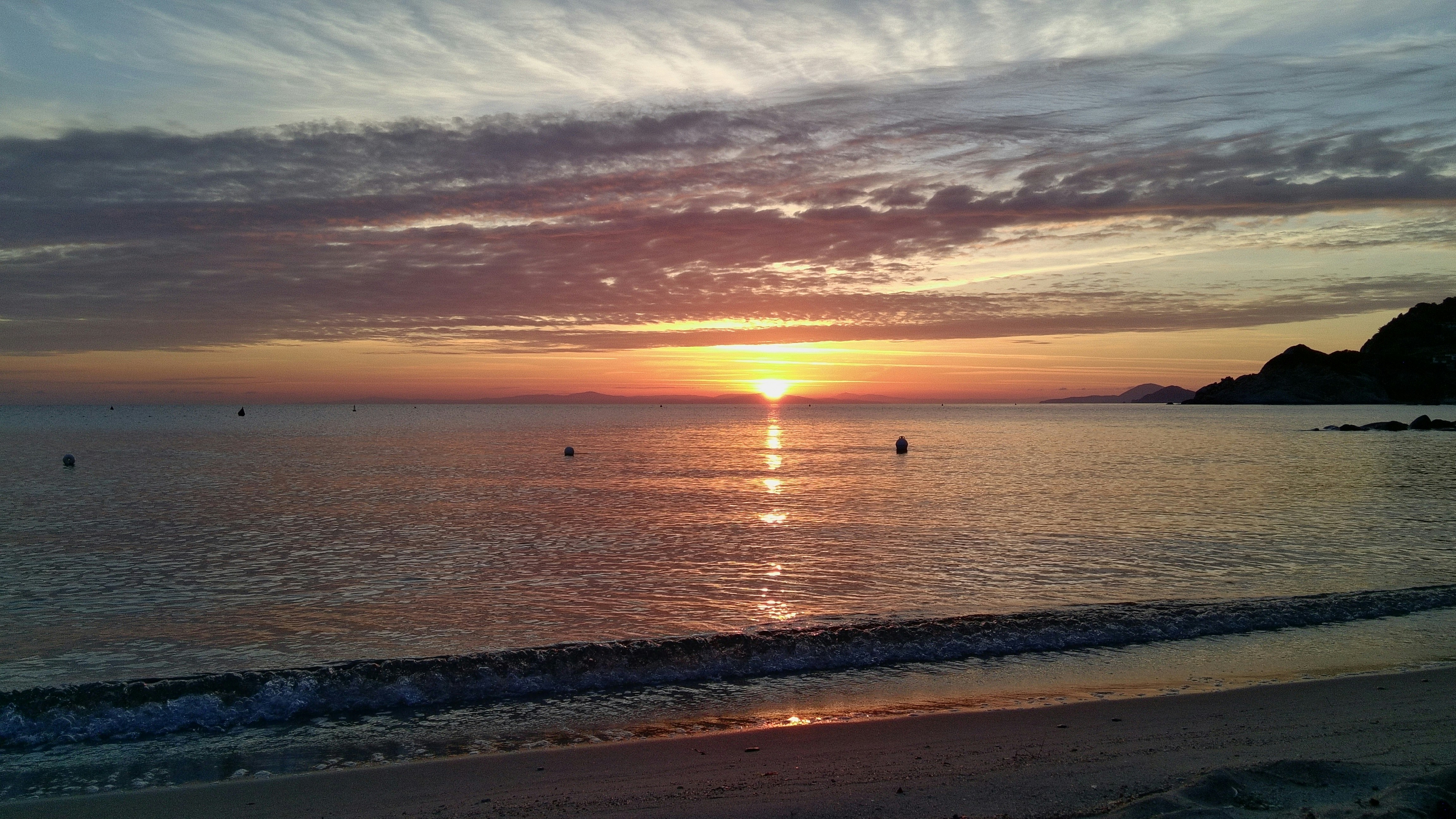Golden sun dipping below the horizon, casting shimmering reflections on the calm sea with gentle waves lapping at the shore.