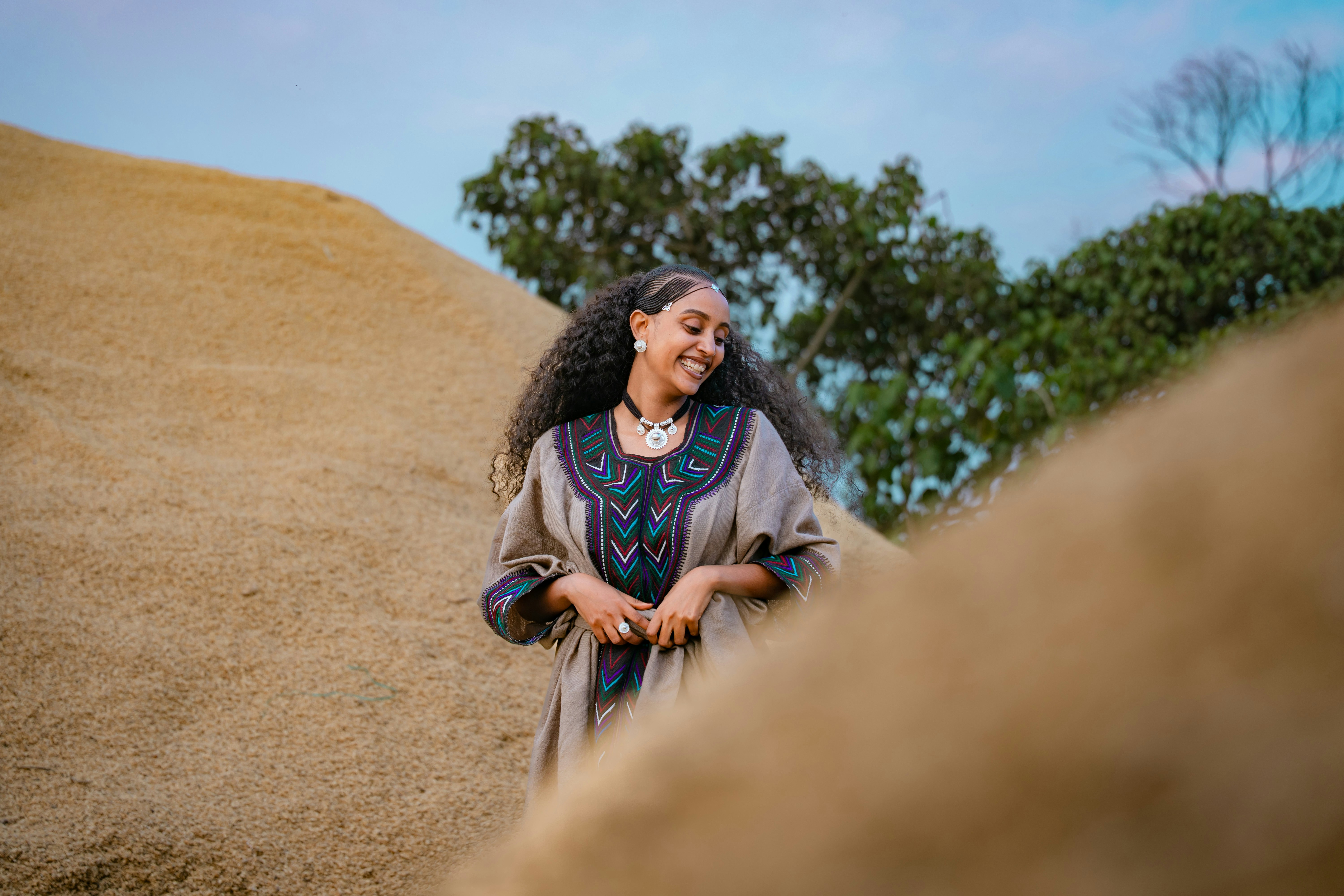 Woman in traditional attire smiles among golden hills, showcasing cultural heritage against a vibrant sky.