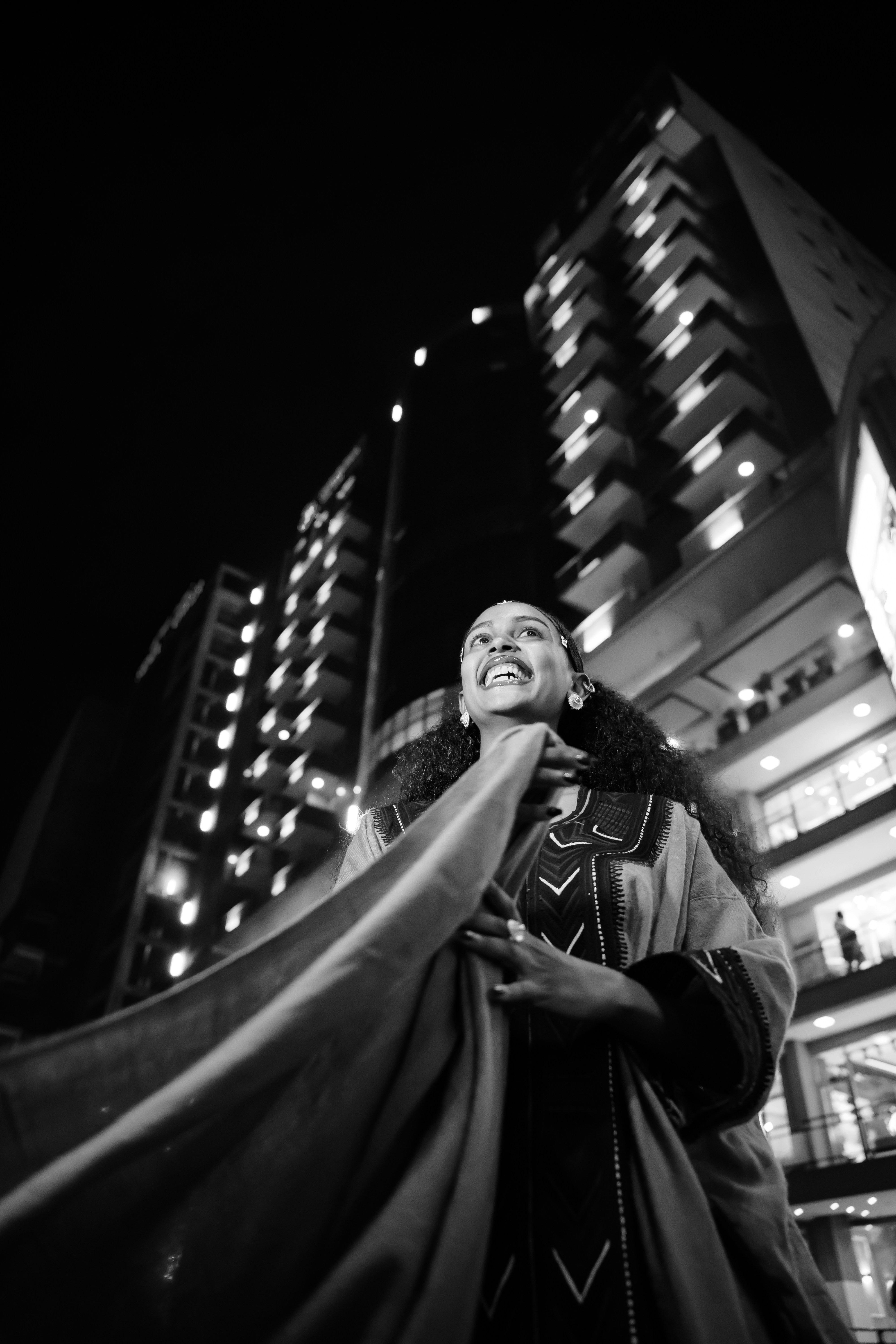 A woman in traditional attire smiles joyfully against a backdrop of modern skyscrapers illuminated at night.