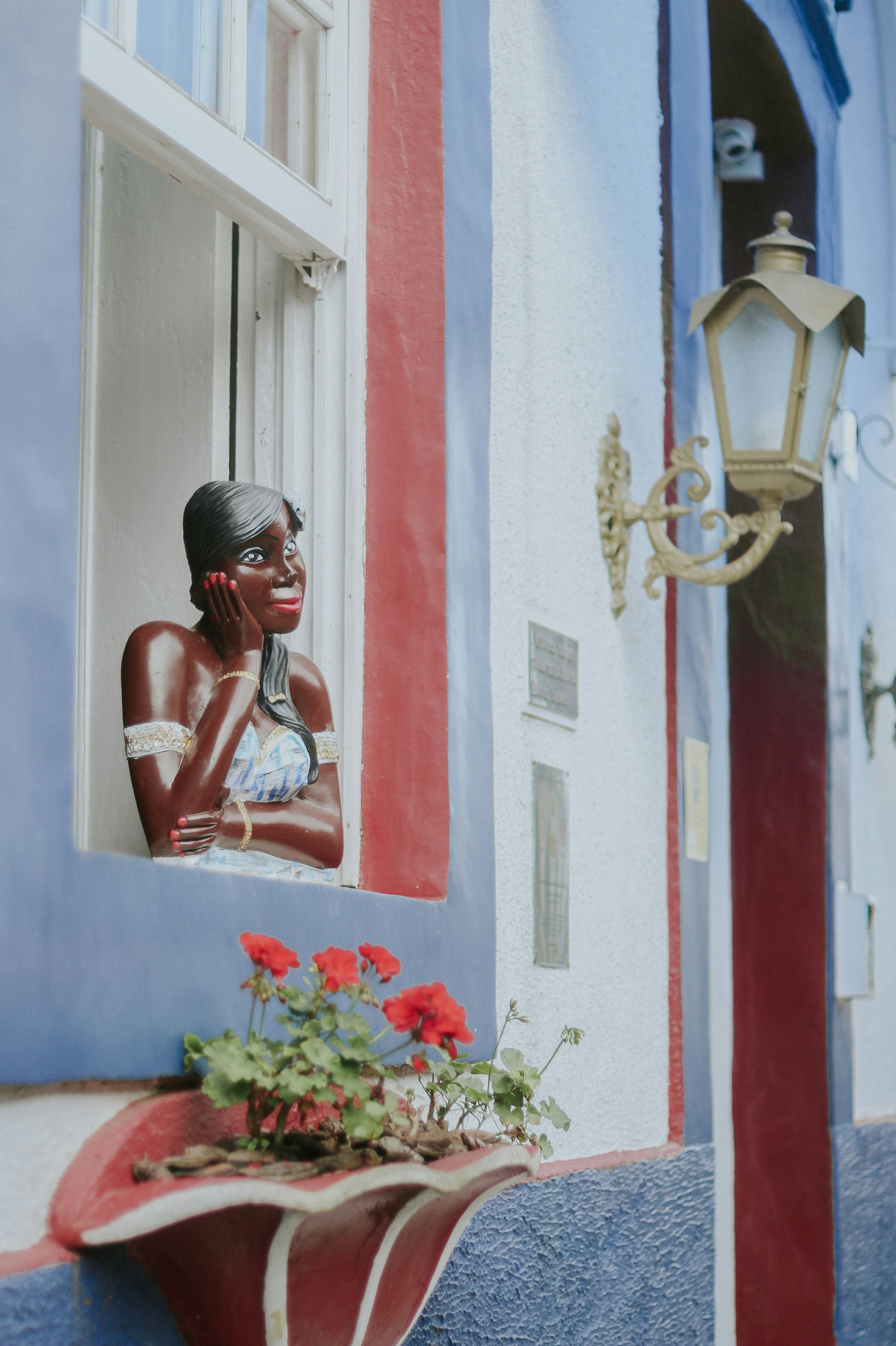 Statue in window with red flowers and lamp.