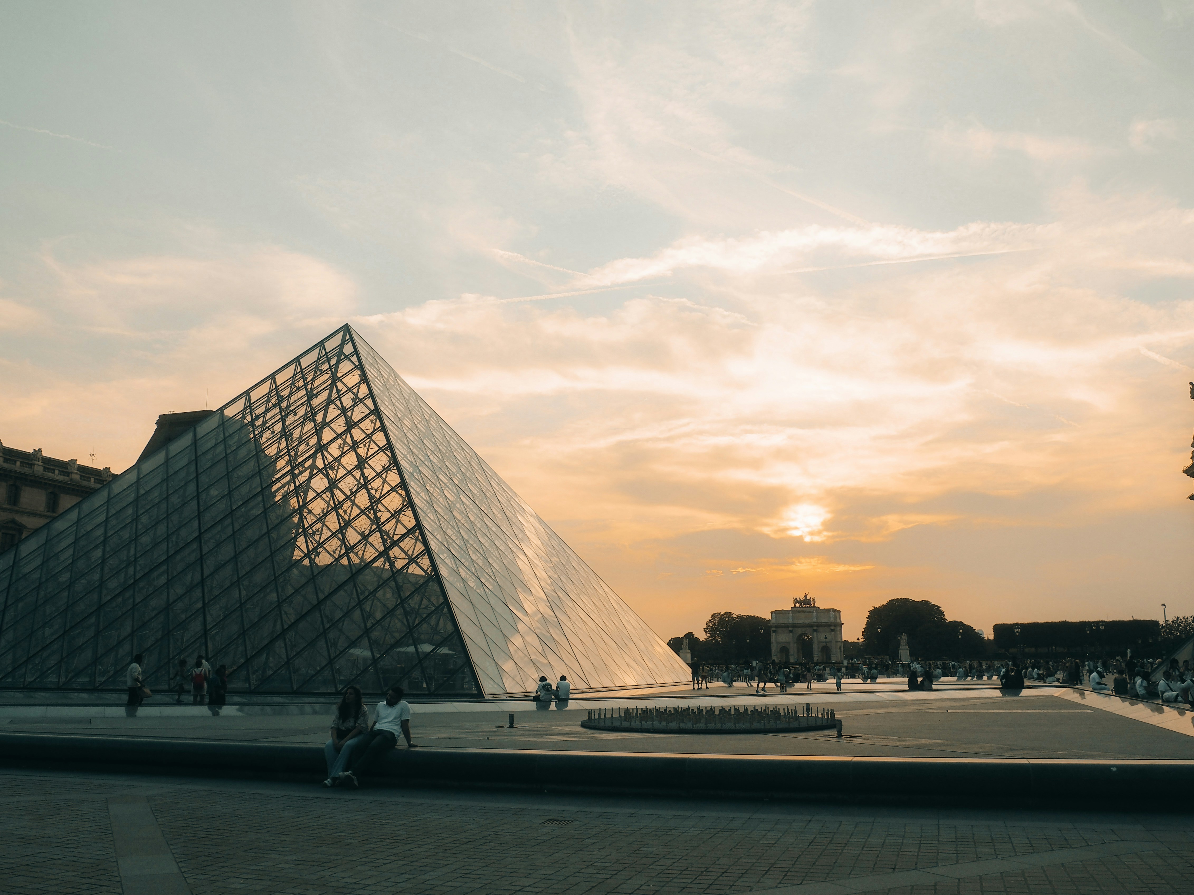 This photo feels like a study in geometry, light, and perspective. The triangular pyramid dominates the composition, its lines pointing upwards while reflecting the soft evening glow. The surrounding palace walls, filled with ornaments, arches, and sculptures, act like elegant frames that emphasize the modern design of the glass pyramid. On the ground, the paved courtyard, the walking silhouettes, and the groups of visitors add a human scale. The pyramid may be monumental architecture, but here it feels approachable, surrounded by the buzz of tourists. It’s a perfect example of how urban phot | Louvre pyramid against a soft sunset sky