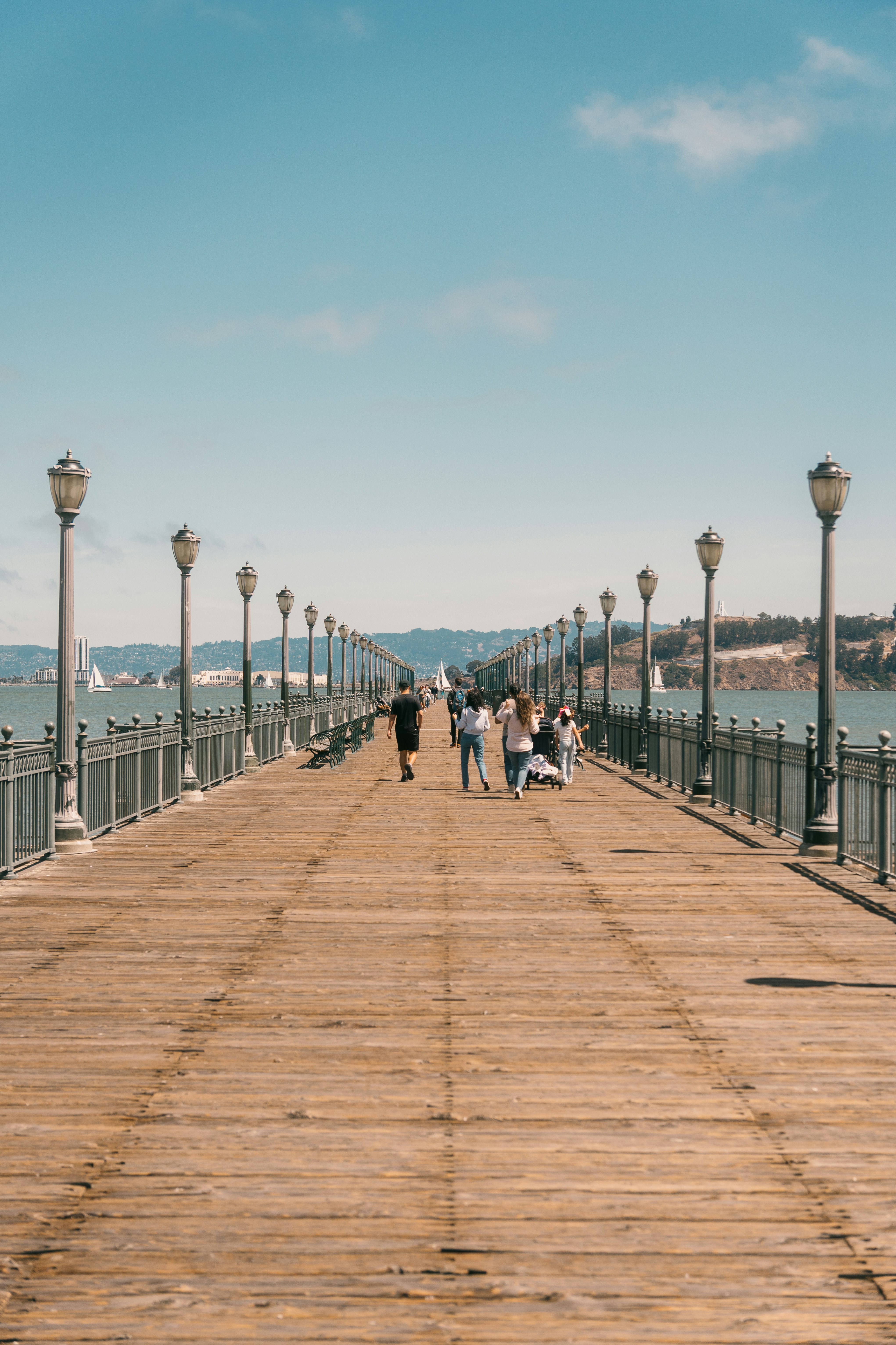 Wooden pier lined with lampposts, bustling with visitors enjoying a sunny day by the water.