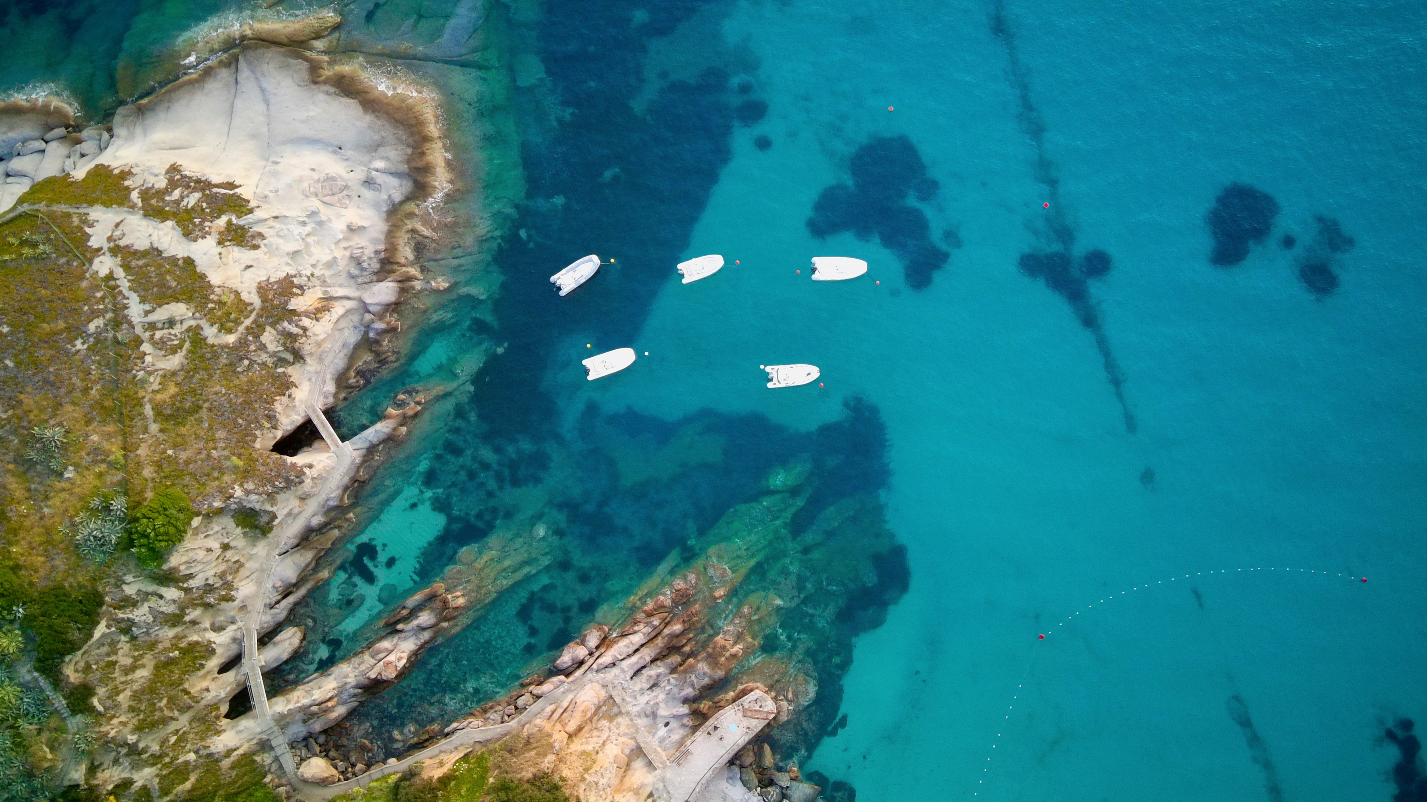 Aerial view of boats anchored in tranquil turquoise waters near rocky coastline, showcasing the harmony between land and sea.
