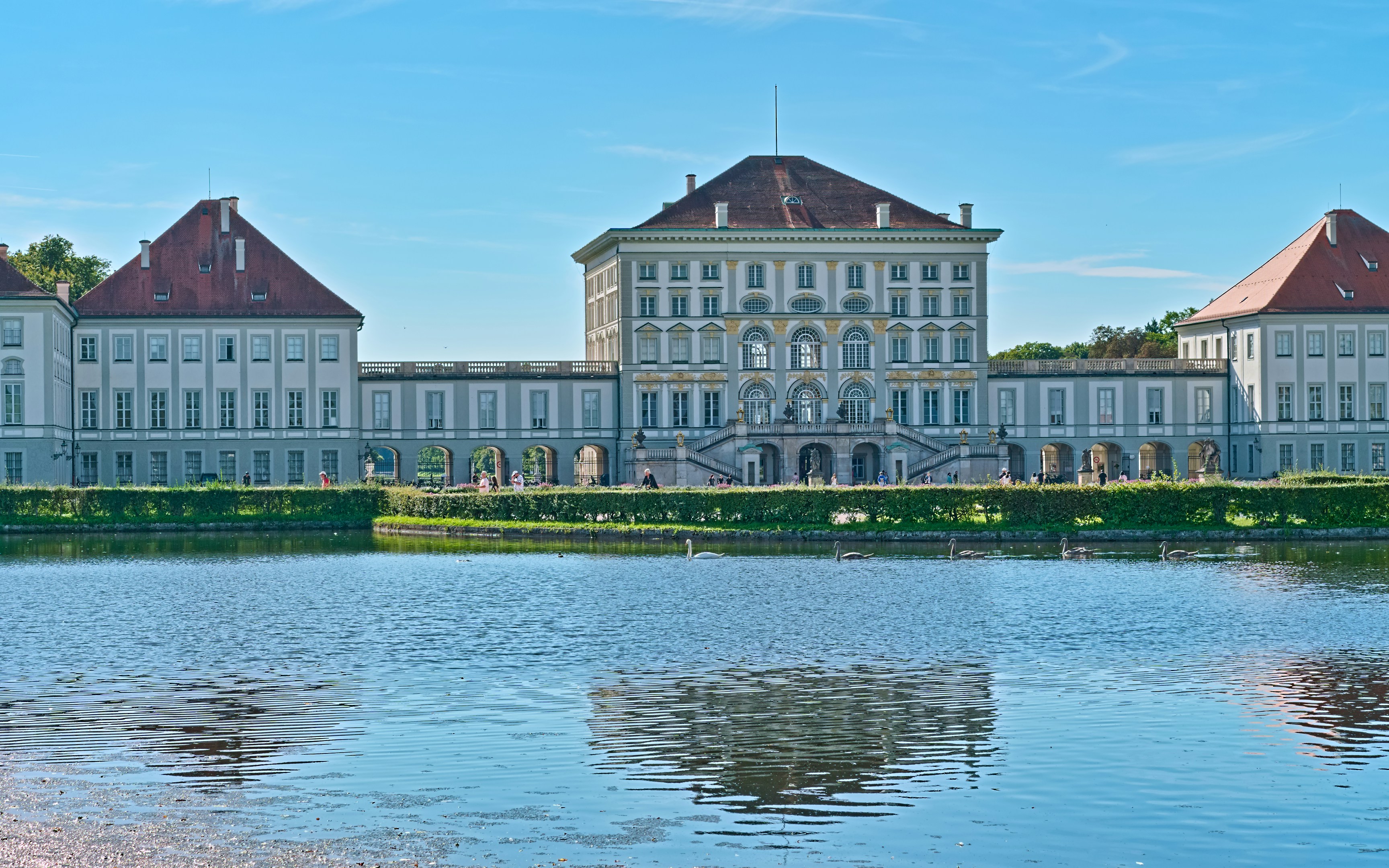 Historic palace reflected in a serene lake, surrounded by manicured gardens and clear blue skies.