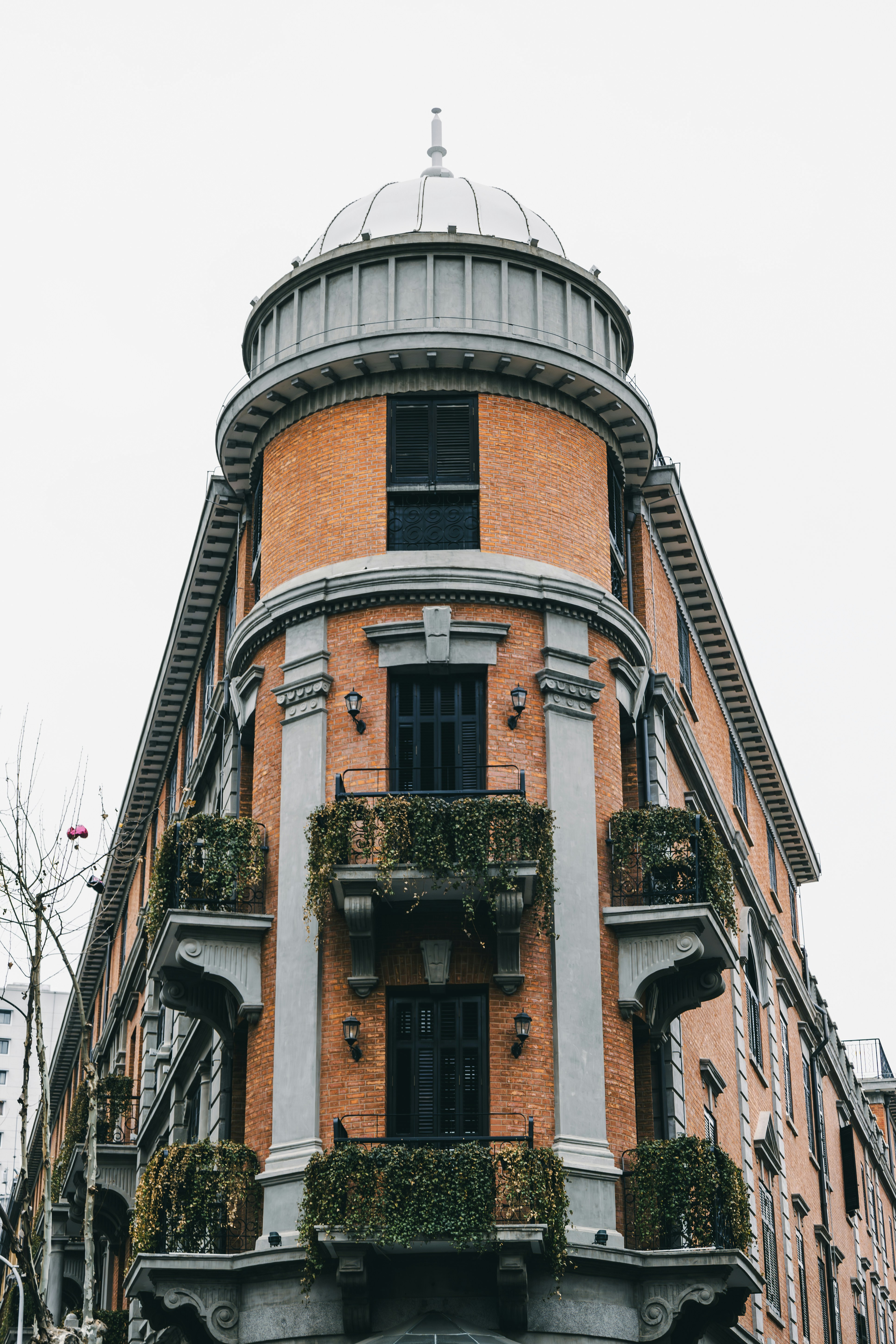 A striking low-angle photograph of a European-style corner building in Wuhan, China. The building features a beautiful facade of warm red brick, contrasted with cool grey stone accents. | Corner building with rounded facade and dome