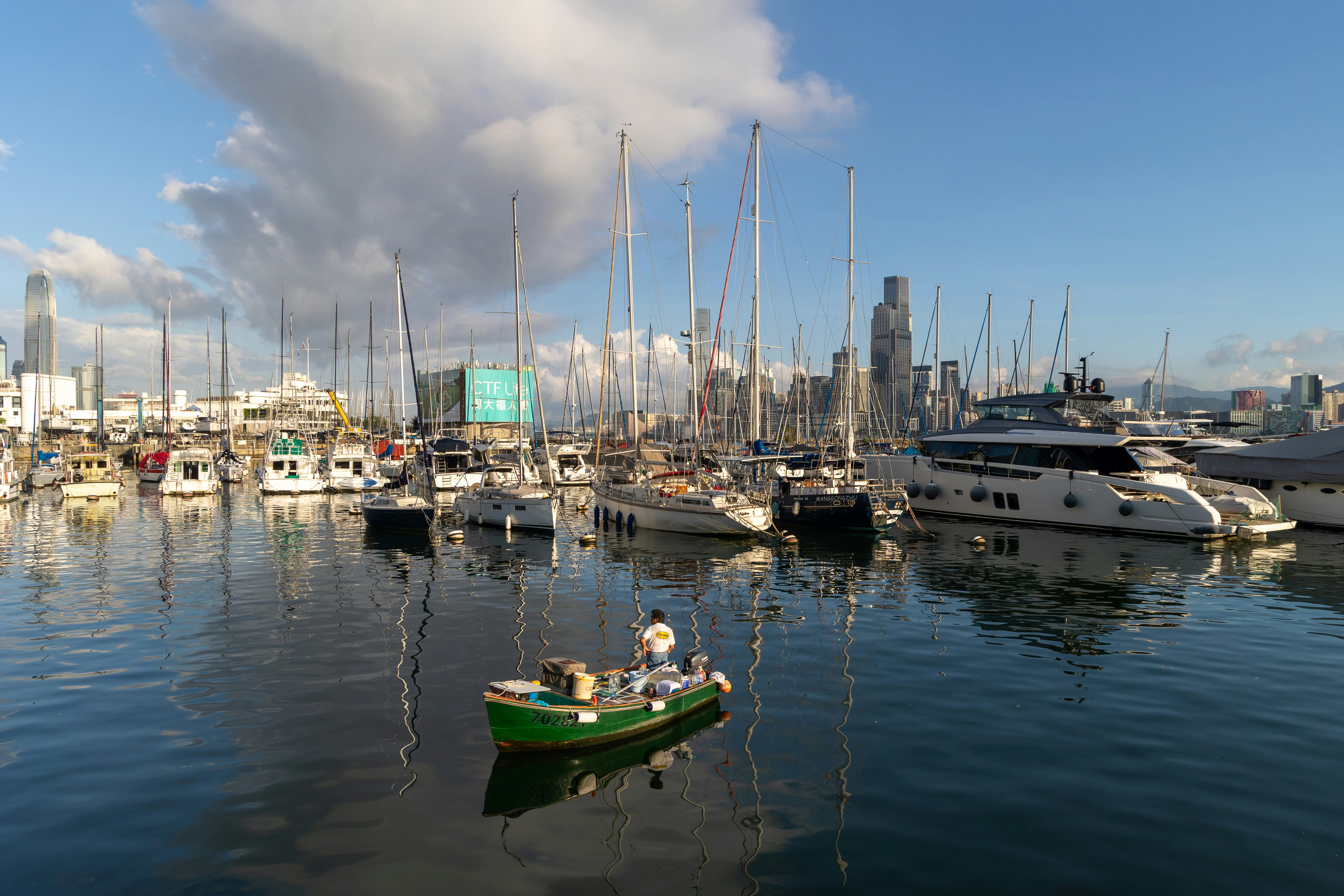 A vibrant harbor scene in Hong Kong with fishing boats and yachts anchored on calm waters. The modern skyline, including iconic skyscrapers, reflects beautifully on the surface, blending urban energy with maritime charm. | Sailboats and yachts docked in a harbor with city skyline.