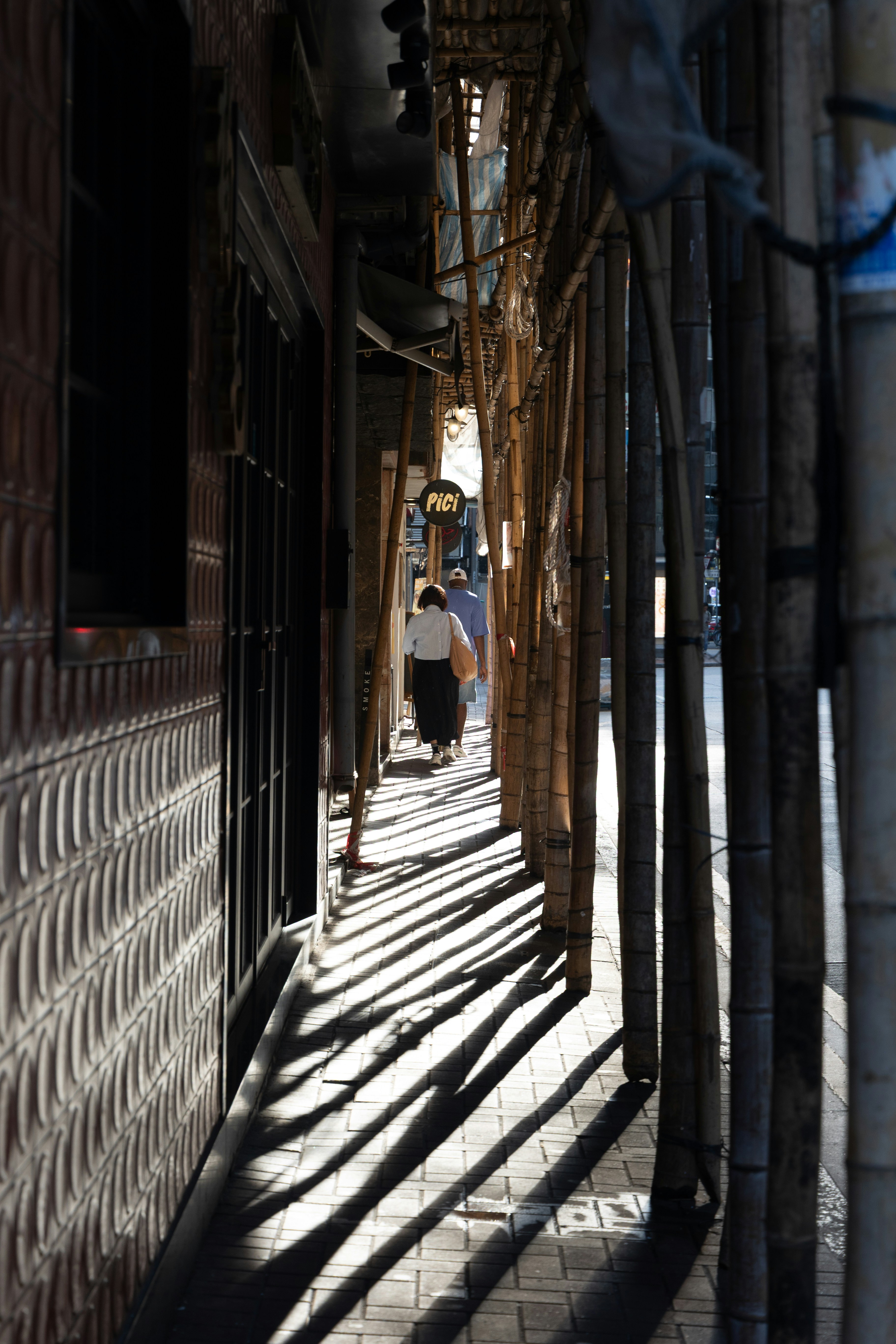 A narrow alleyway in Hong Kong with bamboo scaffolding casting dramatic shadows on the ground. The scene captures the unique urban texture of the city, where tradition and modernity blend in daily street life. | Person walks down sunlit alley with striped shadows.