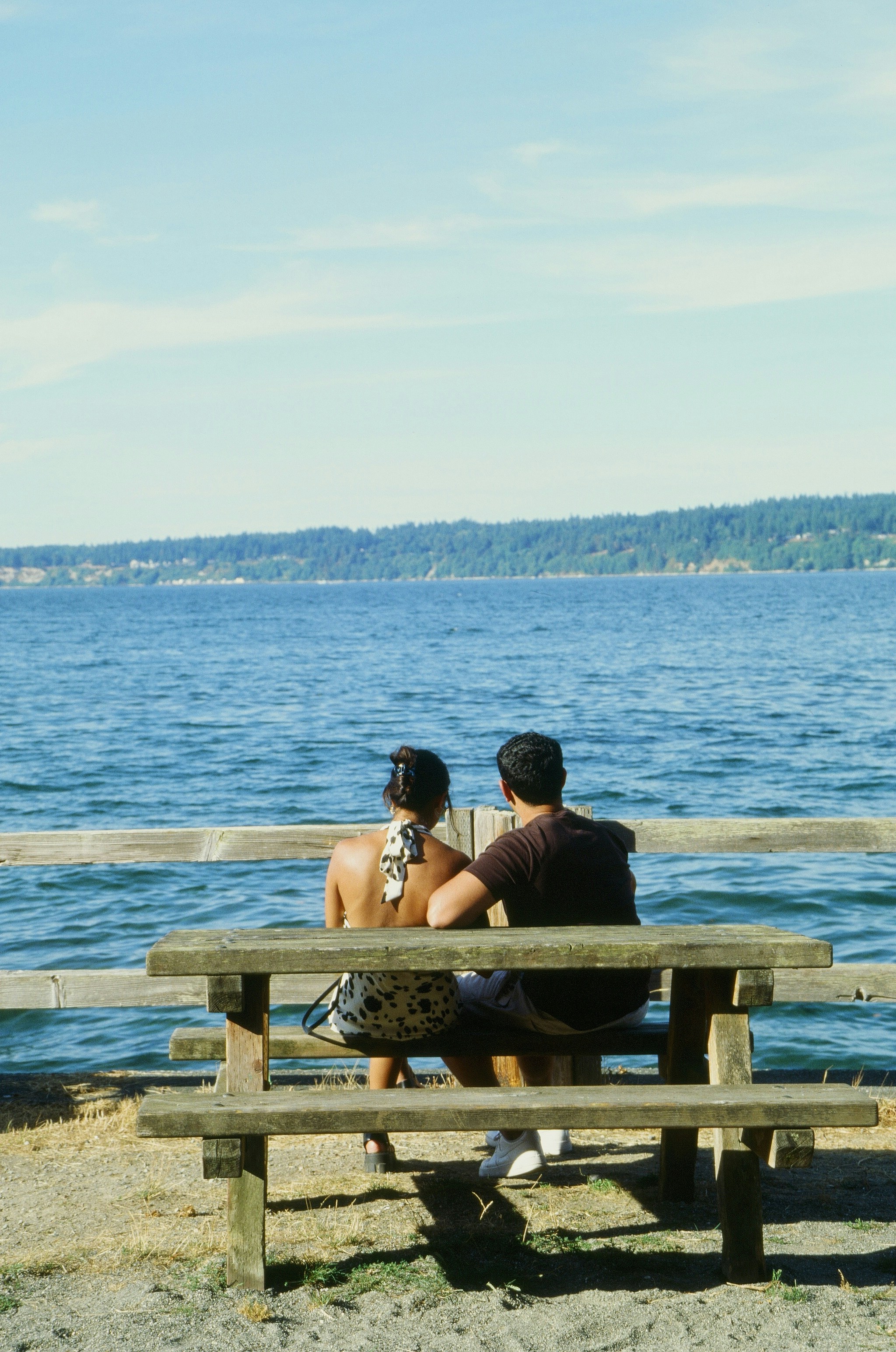 A couple enjoys a peaceful moment by the water, seated on a wooden bench overlooking the tranquil lake under a clear sky.