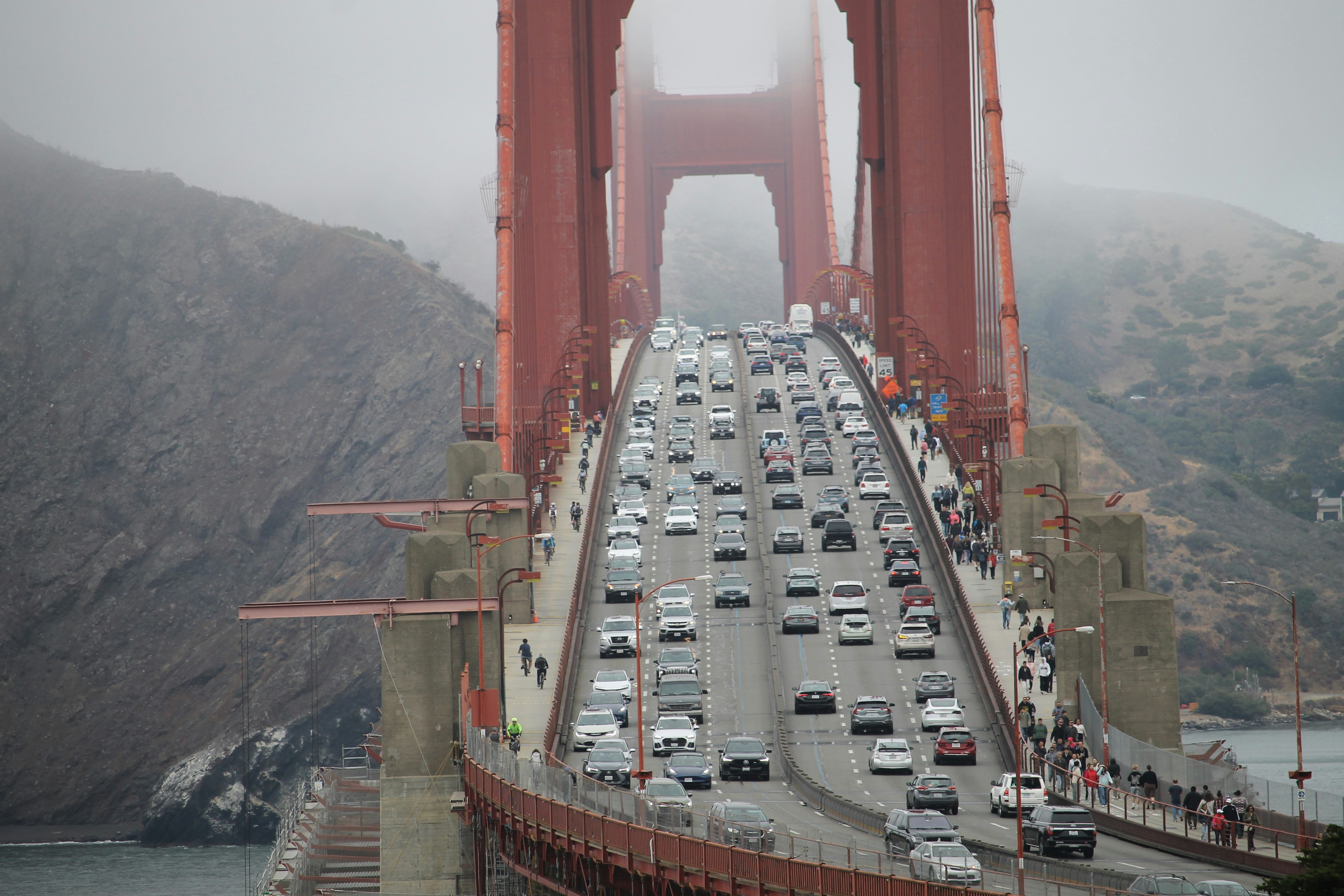 Busy traffic crossing the Golden Gate Bridge, with pedestrians along the walkway and fog enveloping the scene.
