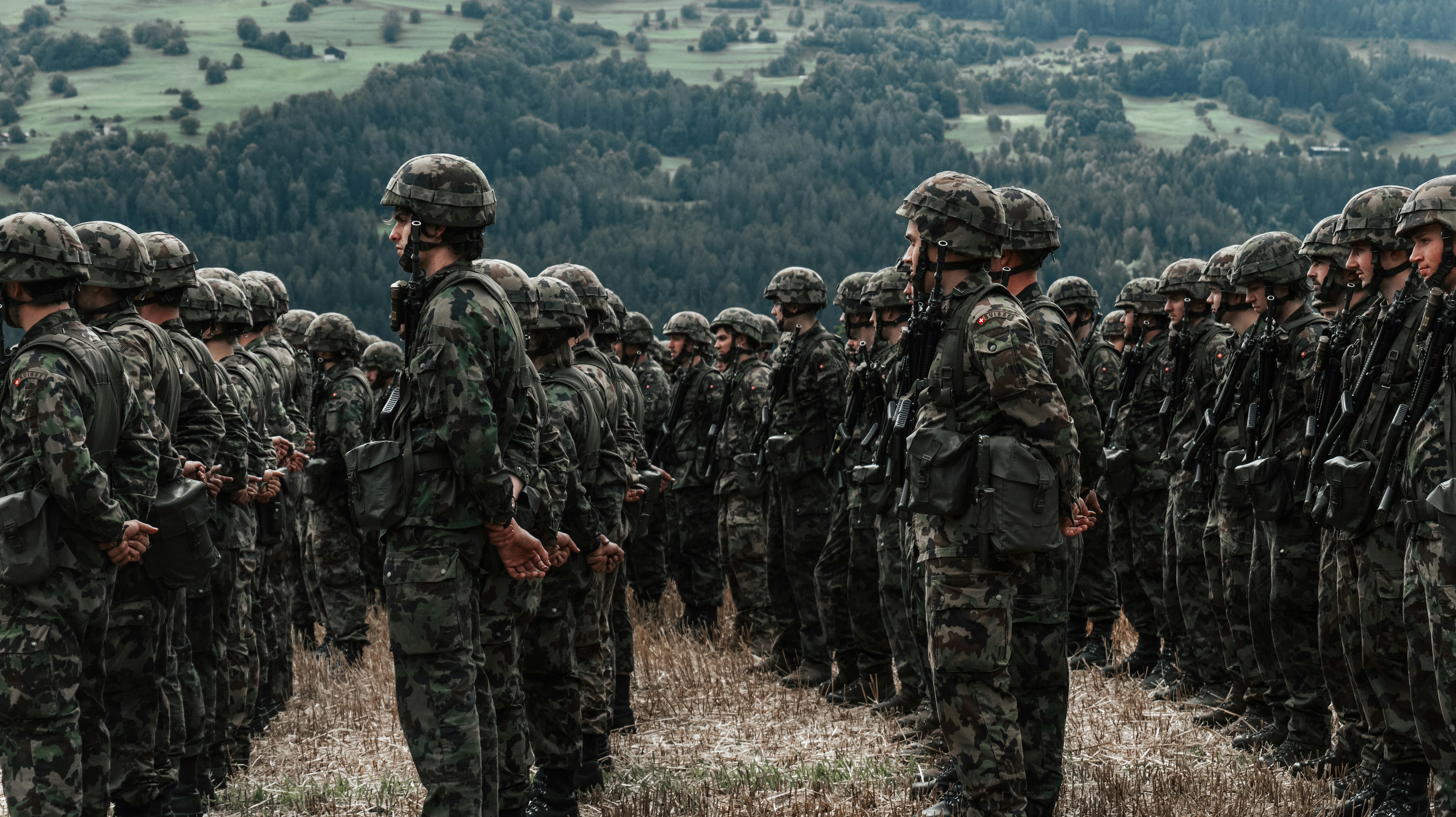 Rows of soldiers in camouflage uniforms stand in formation against a backdrop of rolling hills and trees.