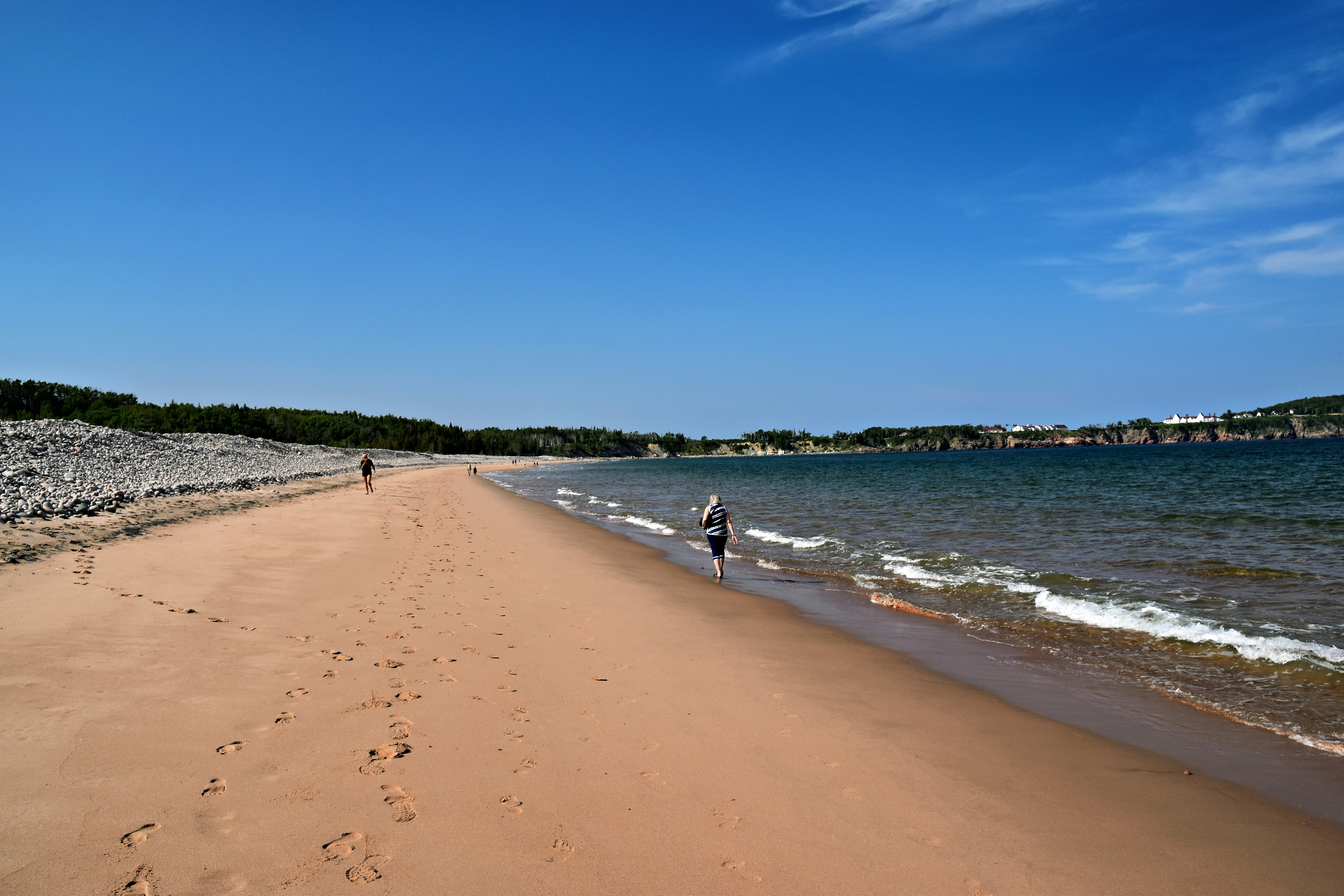 Two figures walking along a sandy beach with gentle waves lapping at the shore under a clear blue sky.