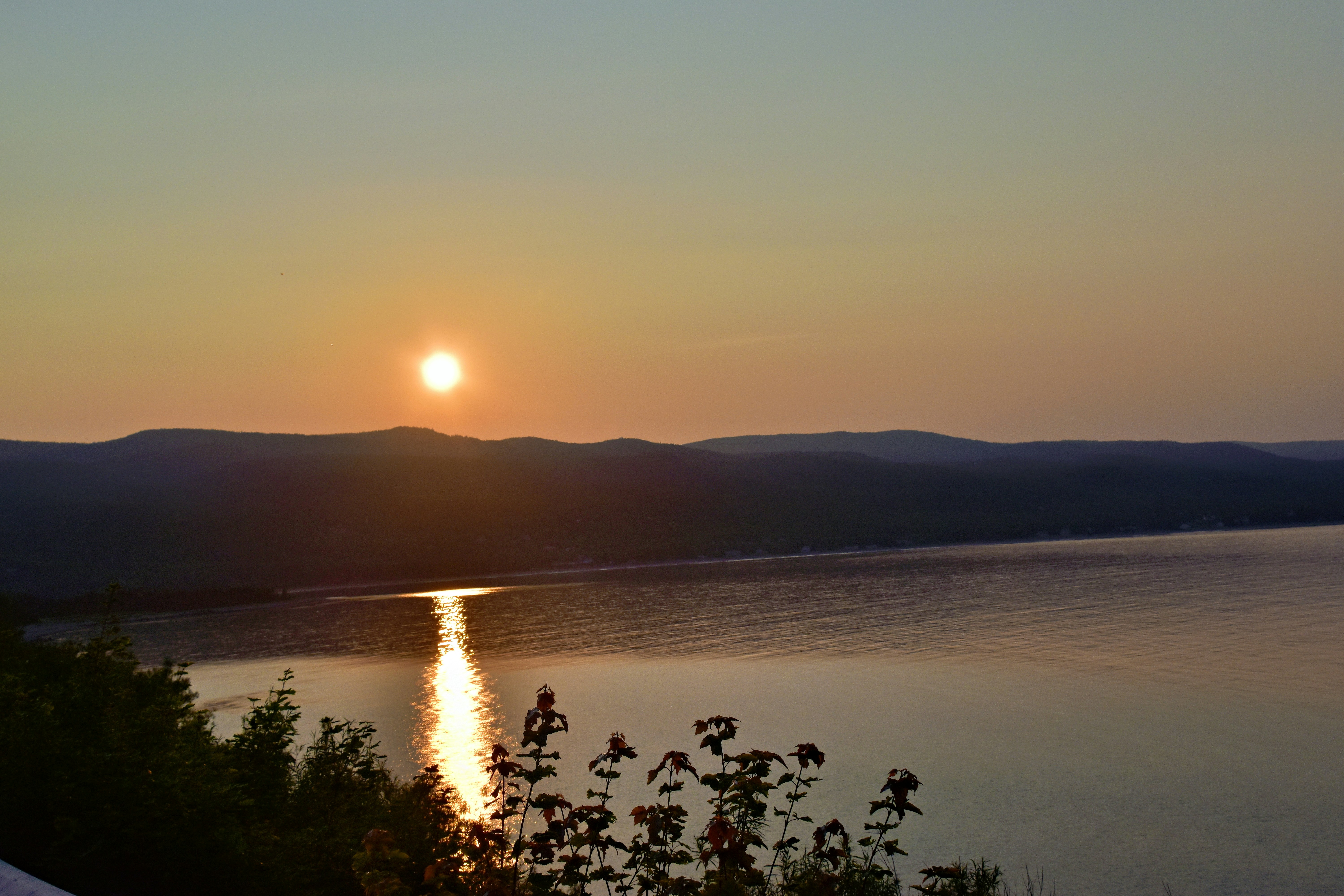 Golden sun setting over tranquil waters, reflecting hues of orange and pink across the landscape. Silhouettes of distant hills frame the scene.