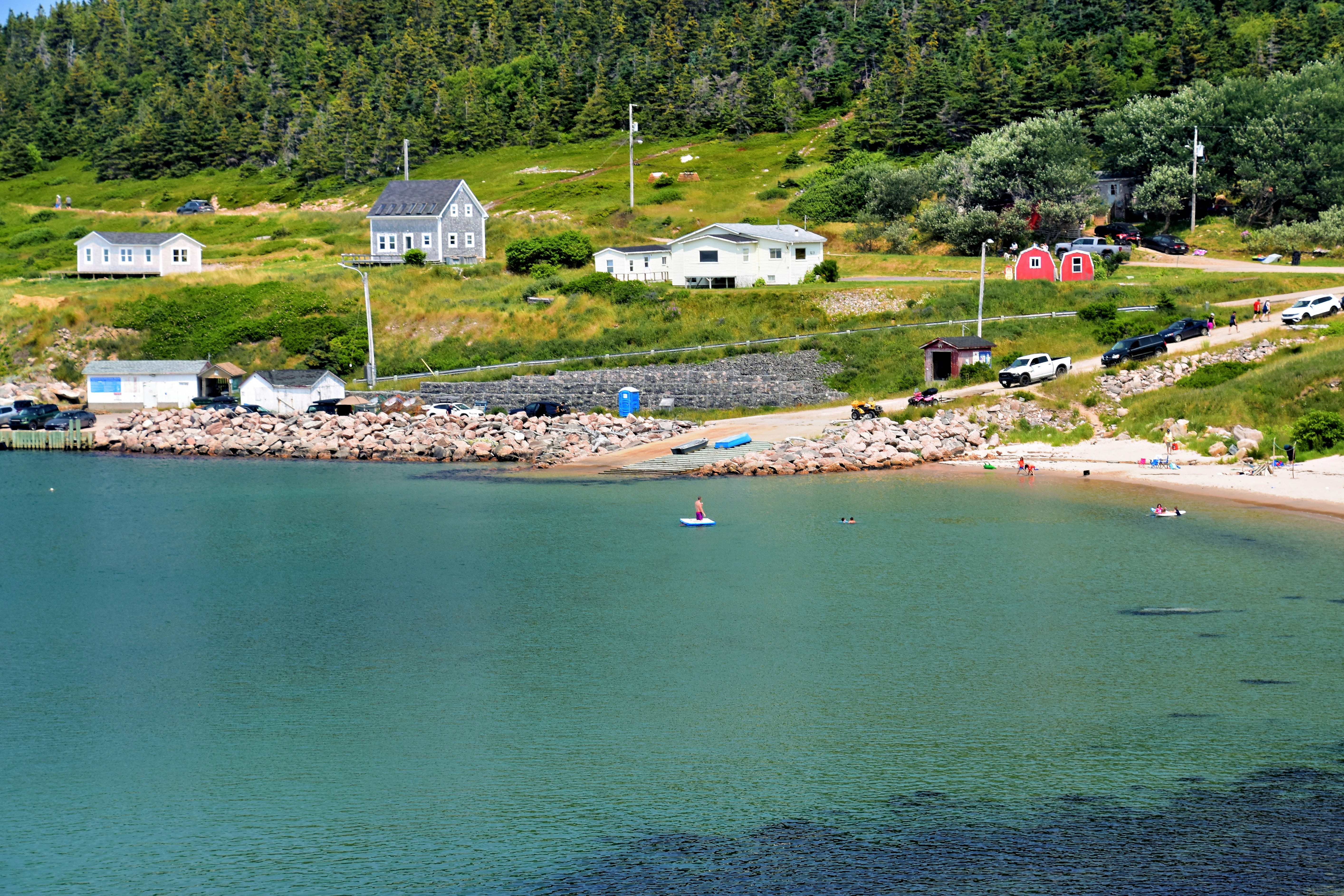 A serene coastal scene showcasing colorful houses along a calm bay, with paddleboarders enjoying the water. The lush greenery in the background enhances the tranquil atmosphere.