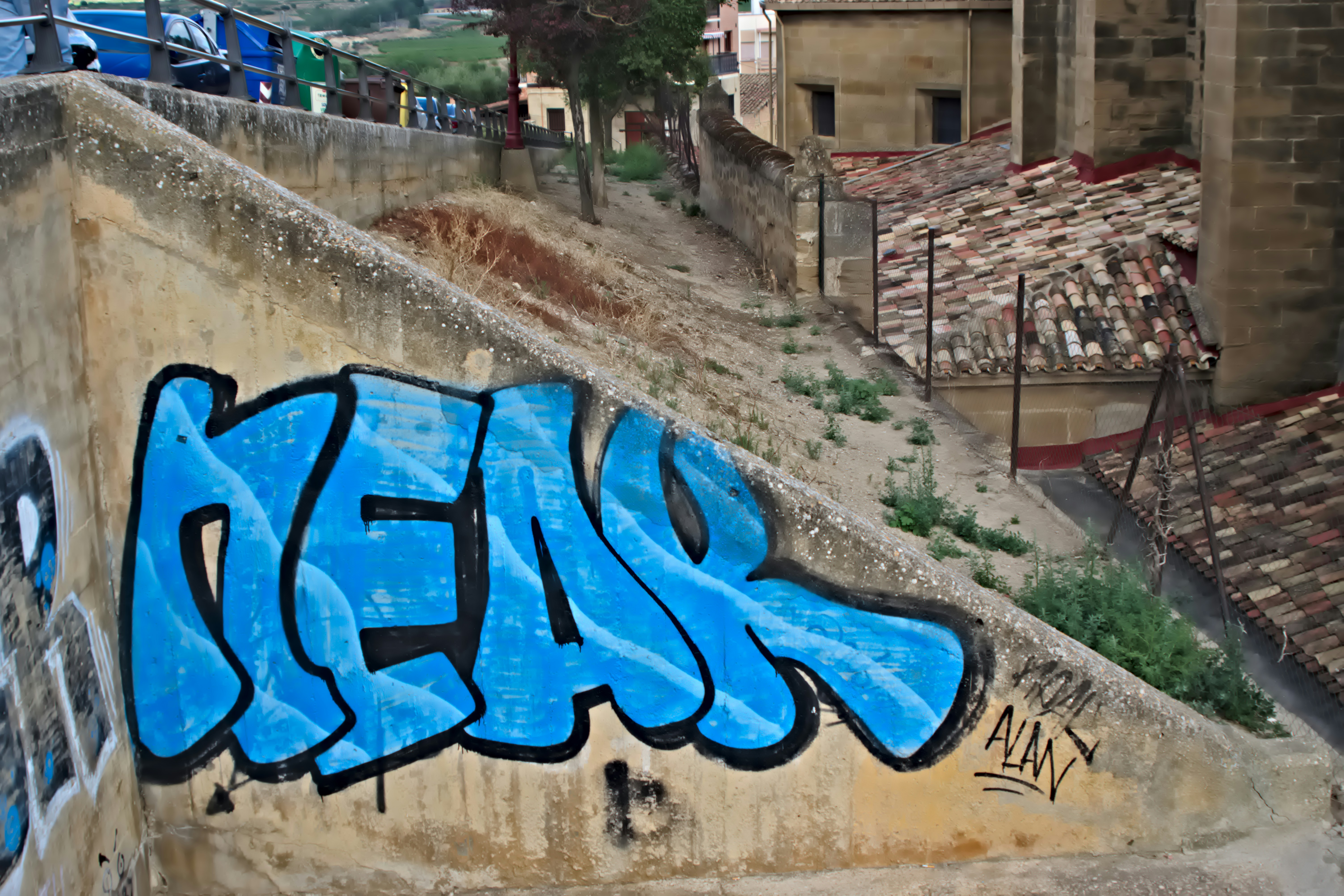 In vittoria, as elsewhere, there are ra many graffiti in the streets | Blue graffiti on a concrete wall with buildings behind