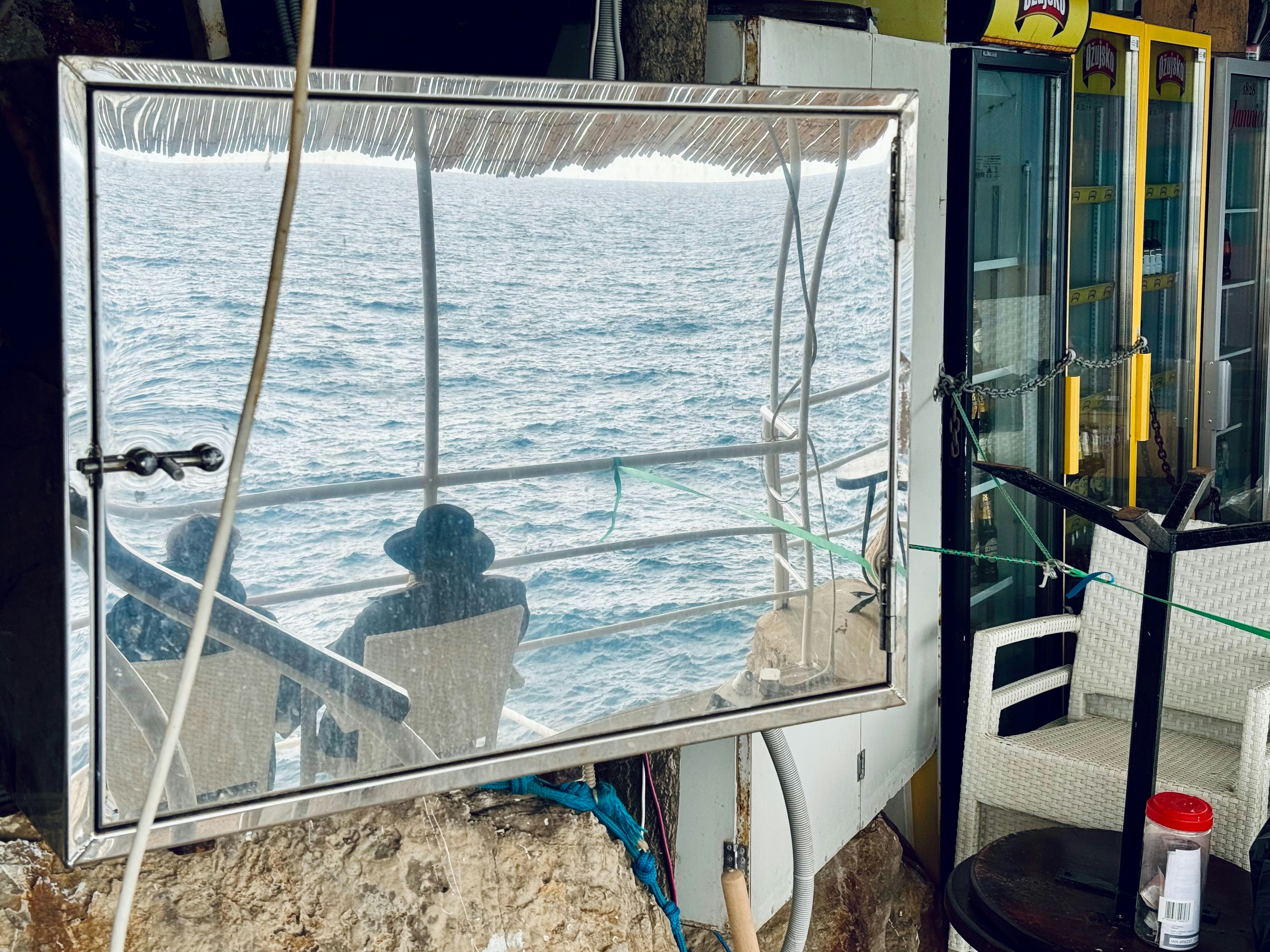 Reflective window showing two figures seated by the seaside, capturing a moment of tranquility and connection with nature.