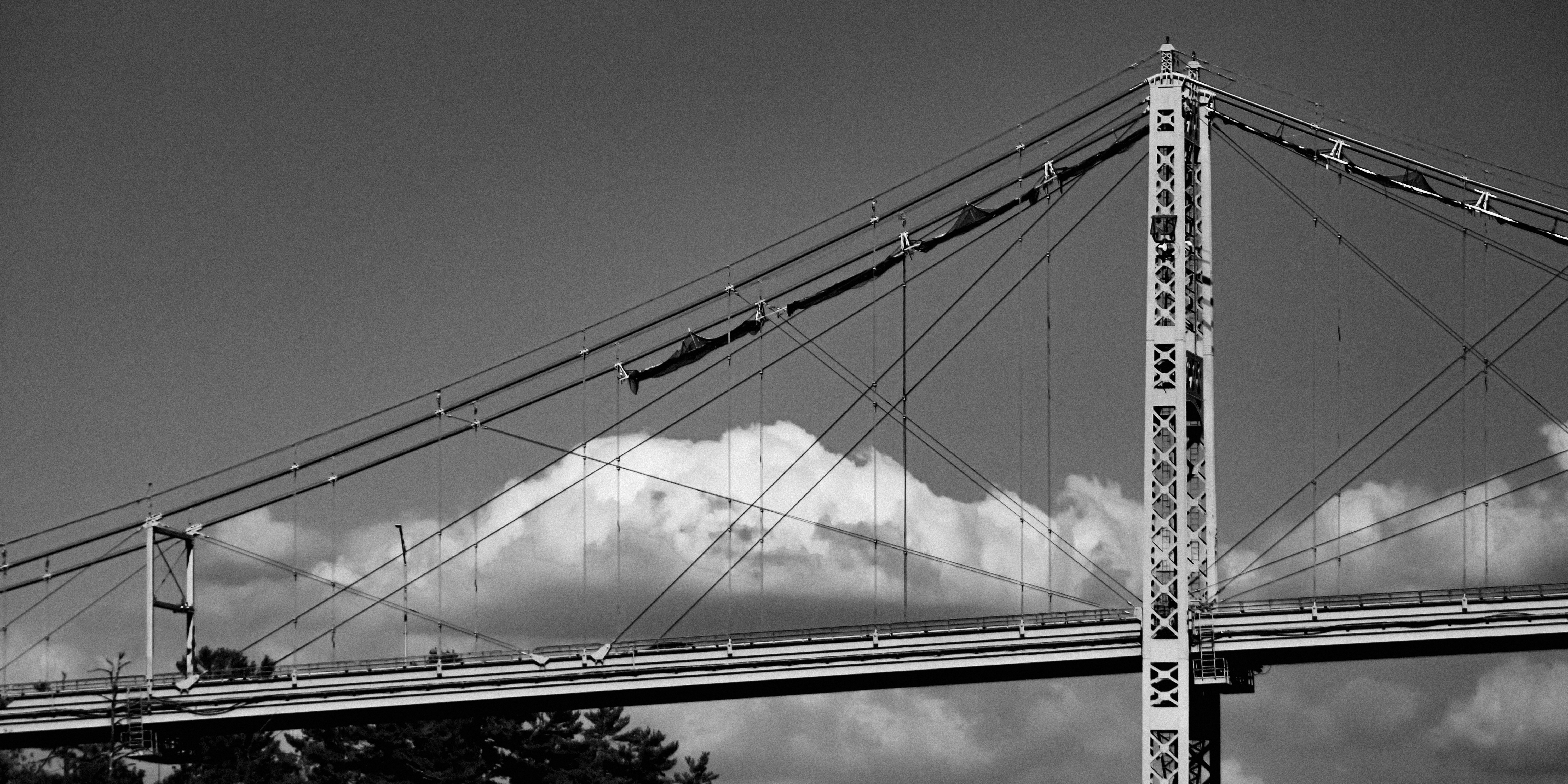 black and white suspension bridge under cloudy sky | Suspension bridge tower with clouds behind it.