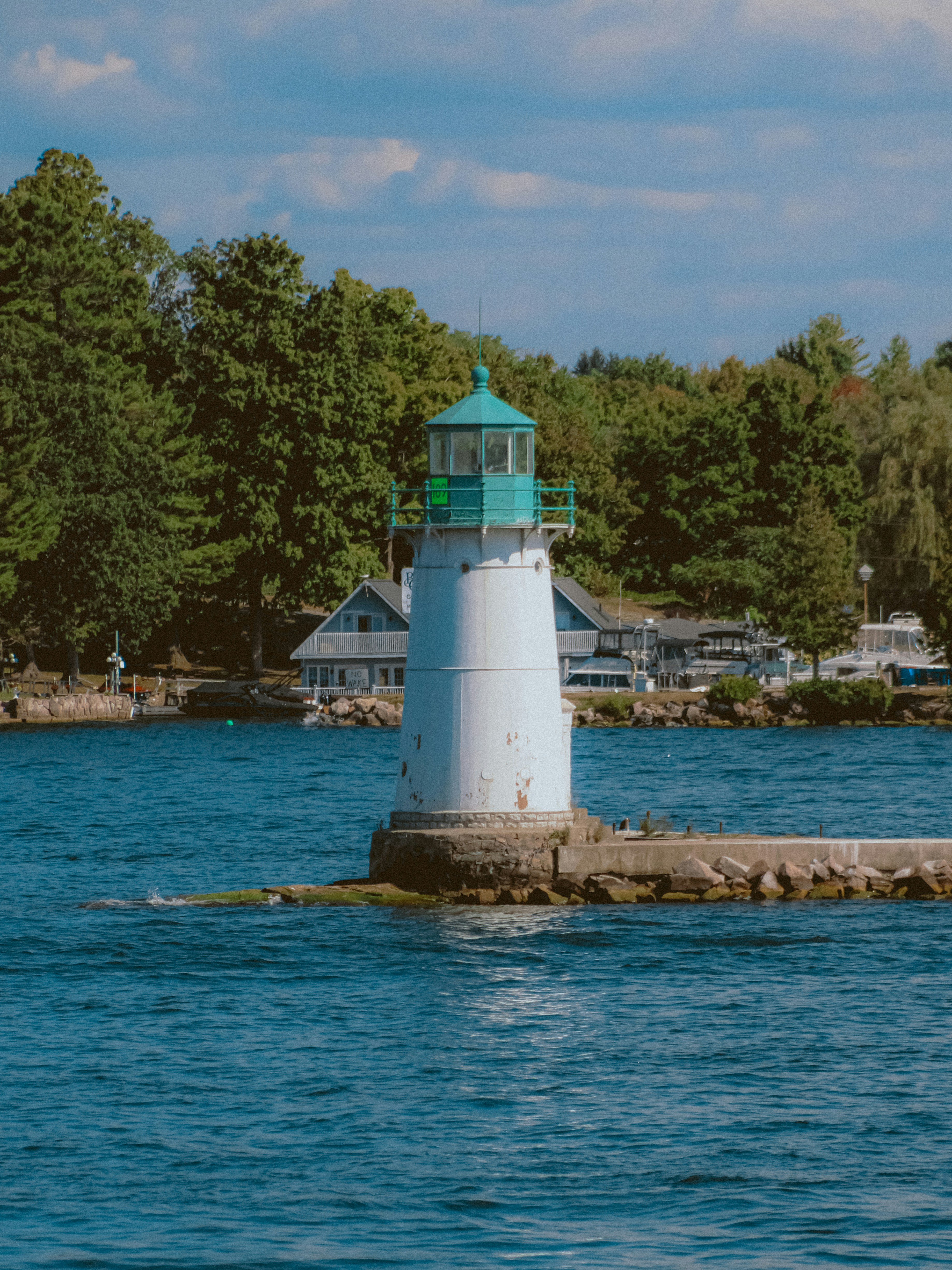 A white lighthouse with a green top stands on a rocky pier, surrounded by lush greenery and calm waters. The serene setting captures the essence of coastal life.