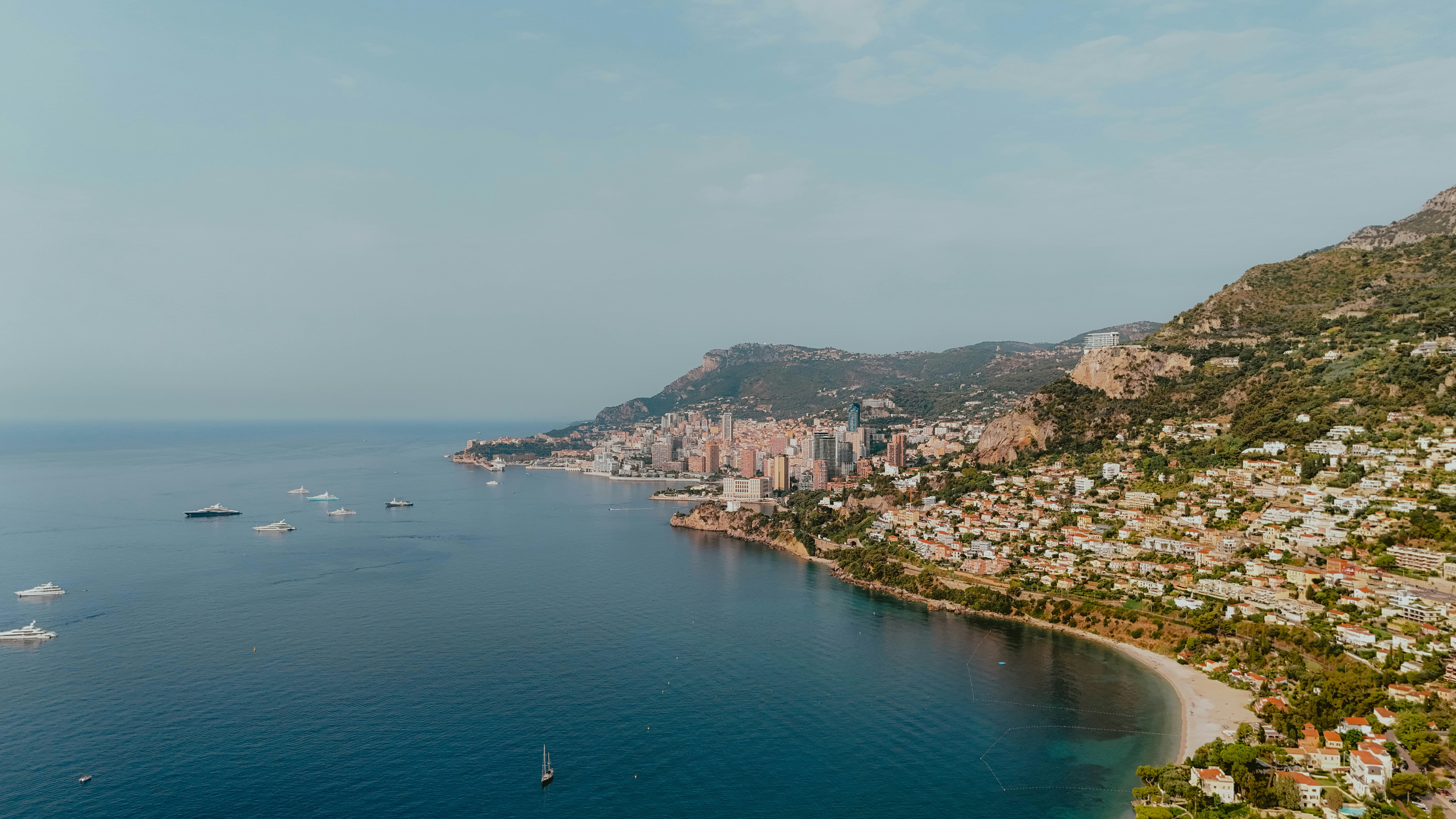 Aerial view of a picturesque coastline with a vibrant city nestled between the mountains and the sea, showcasing boats dotting the tranquil waters.