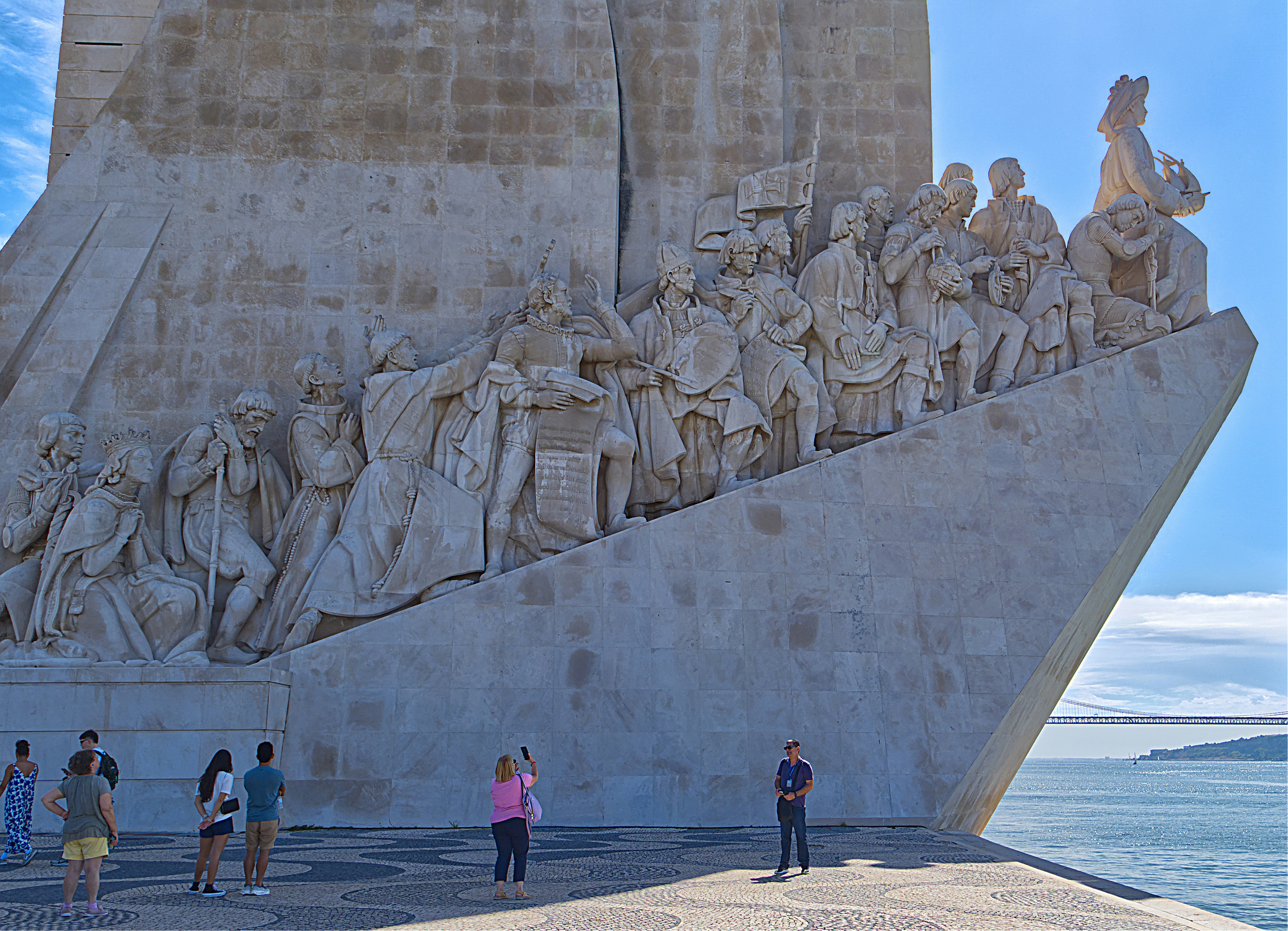 Intricate stone carvings of historical figures celebrate maritime exploration, with visitors admiring the monument against a serene waterfront backdrop.