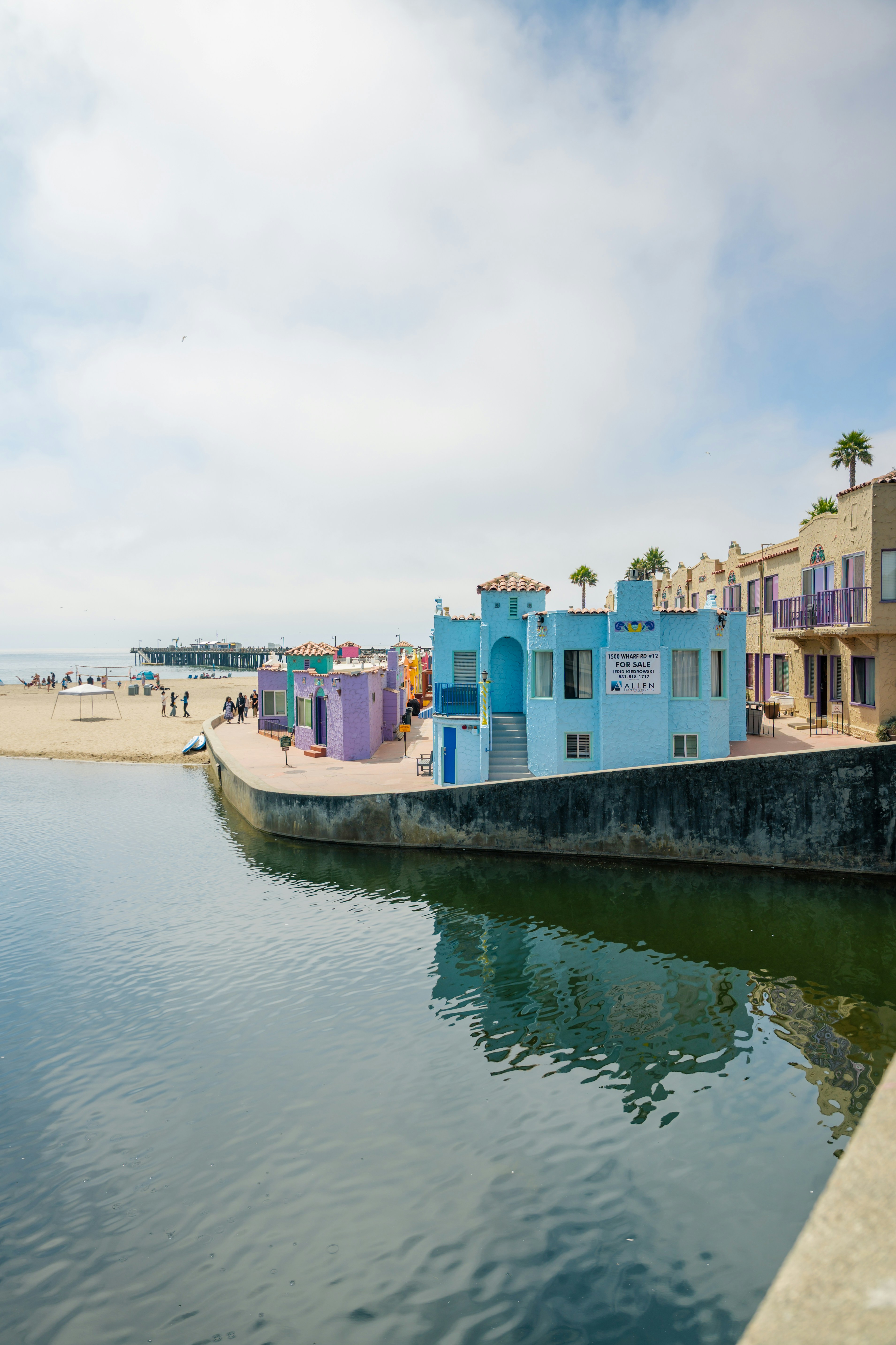 Colorful beachfront buildings reflecting in calm waters, with a sandy shore and distant pier. A tranquil coastal scene unfolds.