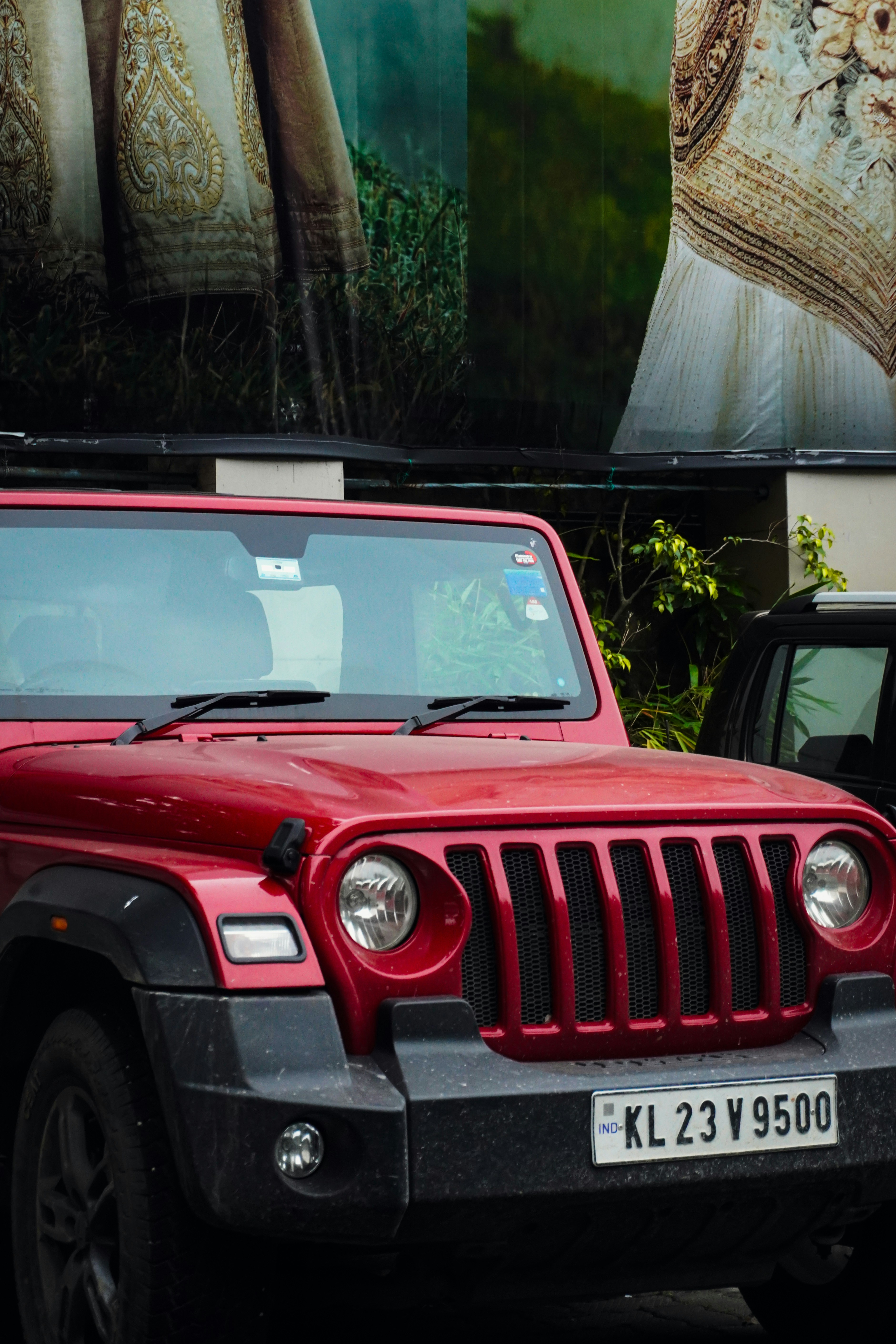A vibrant red jeep parked in front of a large banner showcasing intricate traditional attire, highlighting the juxtaposition of modern and cultural elements.