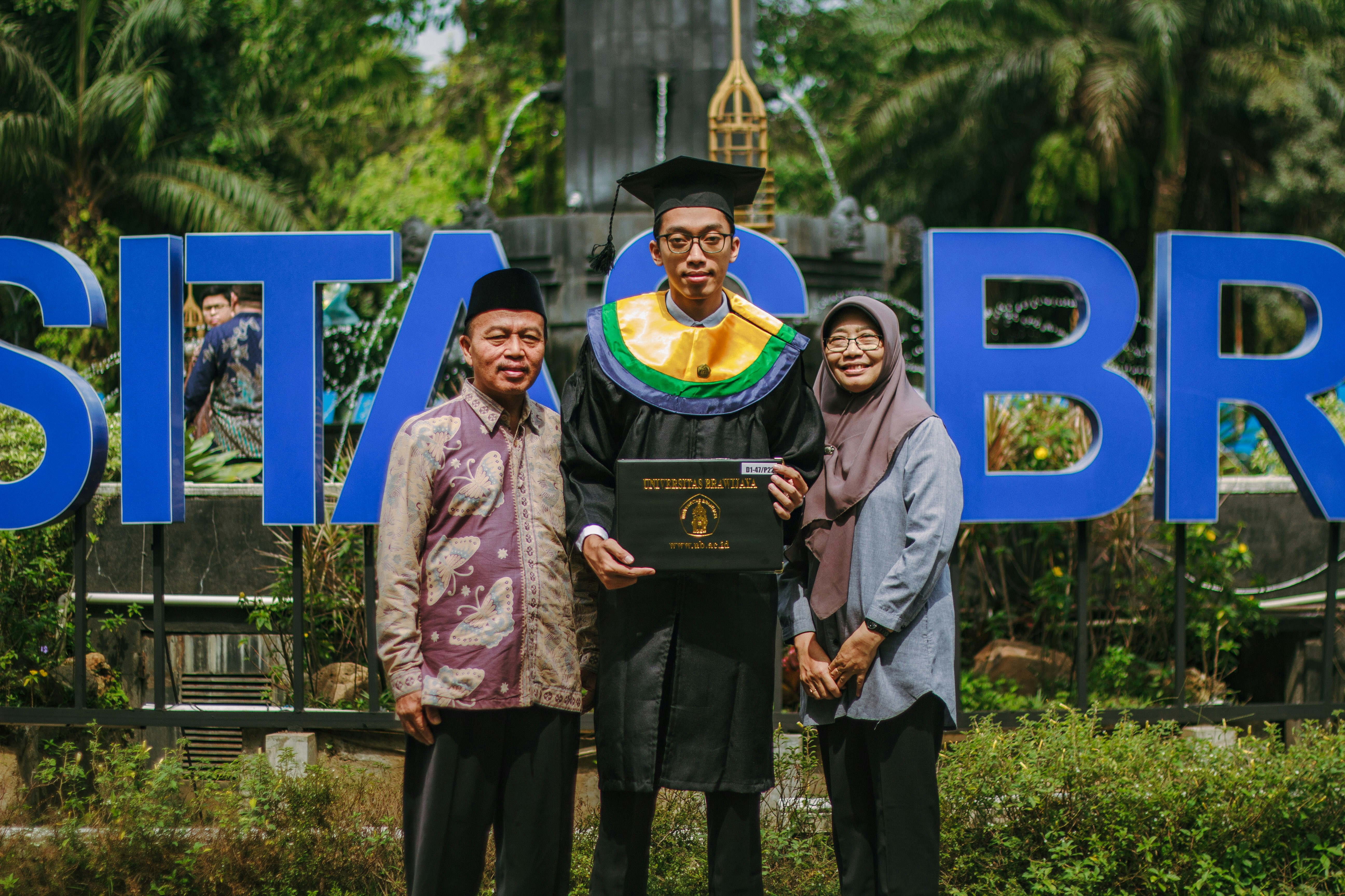 Graduating student proudly holds diploma with family members against a vibrant university backdrop. 