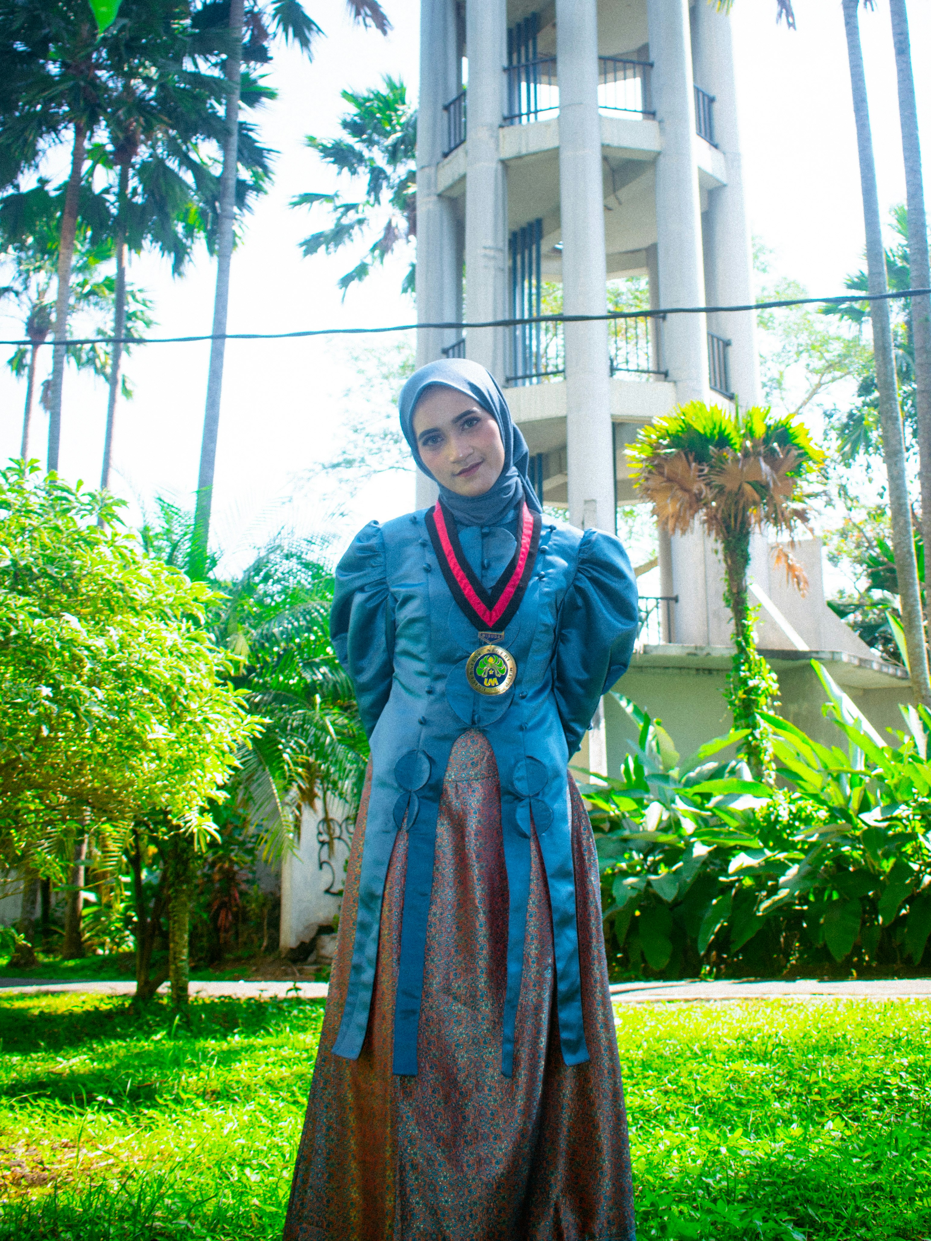 A young woman in traditional attire stands confidently in a lush garden, framed by tall palms and a modern tower in the background.