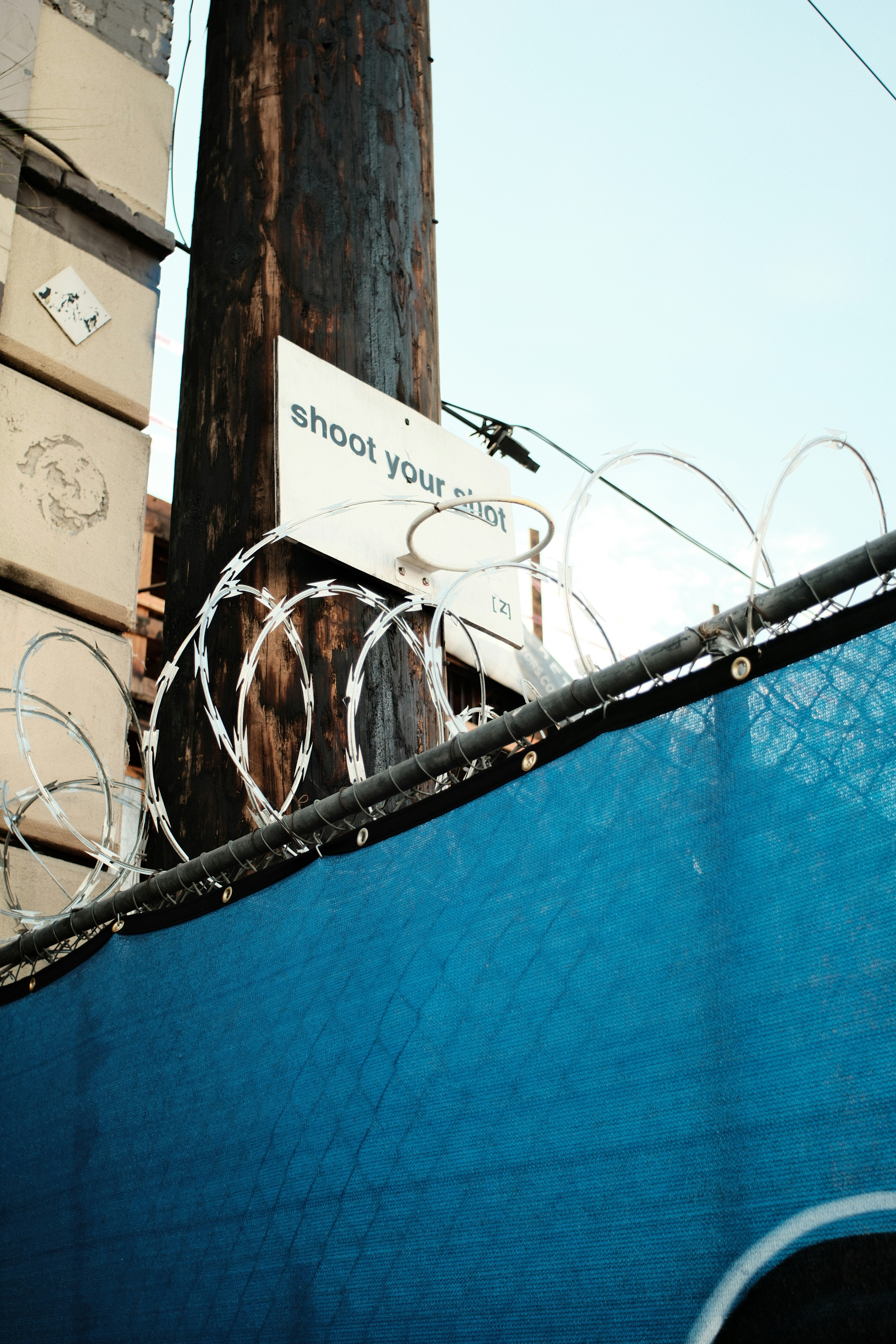 A weathered sign reading 'shoot your shot' mounted on a utility pole, surrounded by barbed wire and a blue construction barrier.