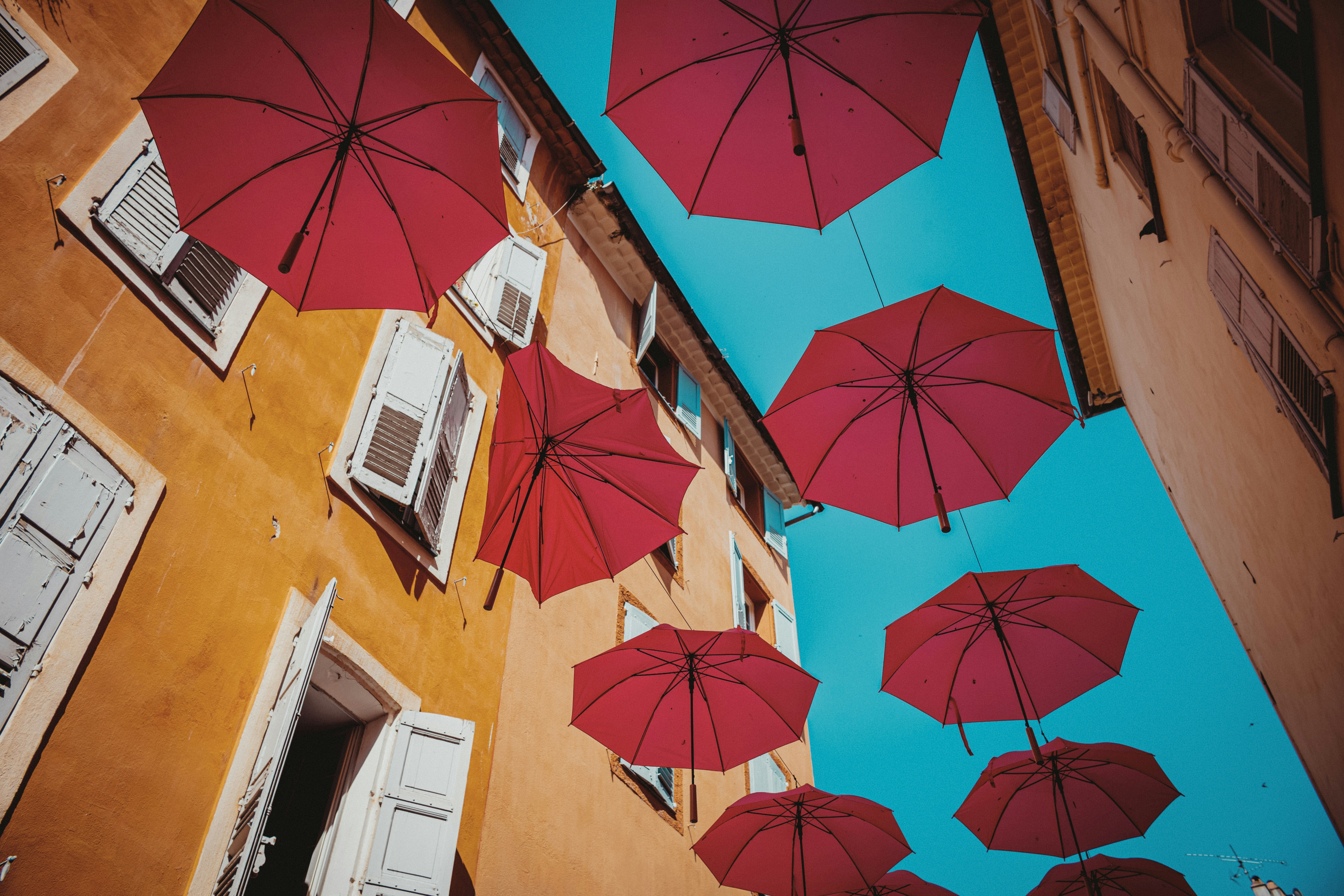 Vibrant red umbrellas suspended above a sunlit street with warm yellow walls and white shutters.