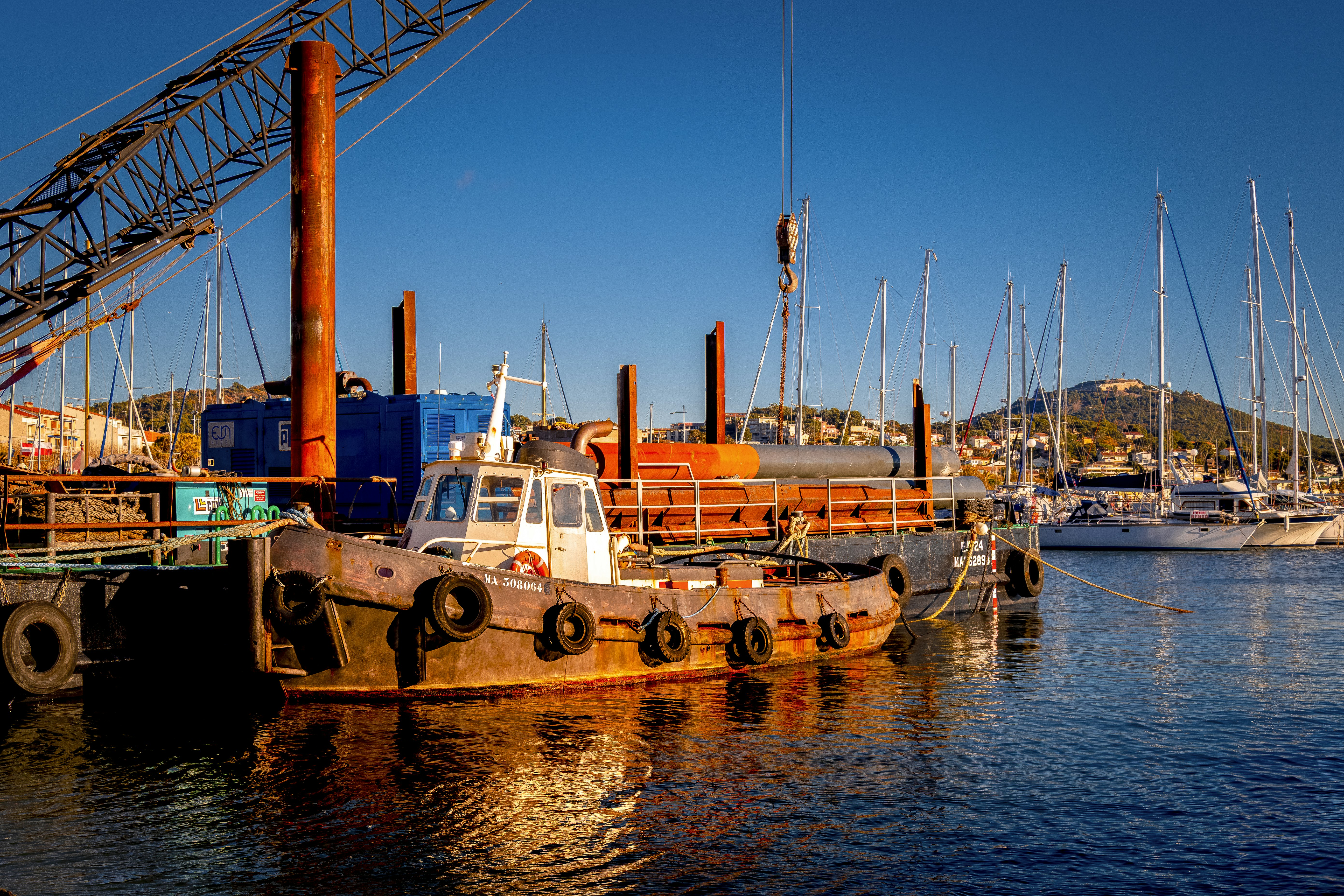 Rusty tugboat docked in a sunny harbor with sailboats.