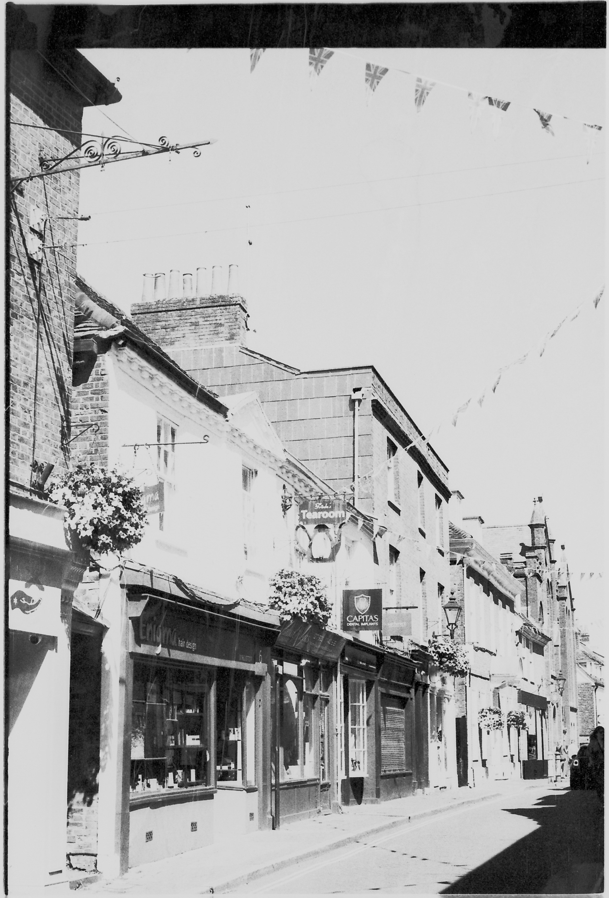 Quaint street lined with historic buildings, adorned with flowering planters and festive bunting. A tea room sign adds a touch of local charm.