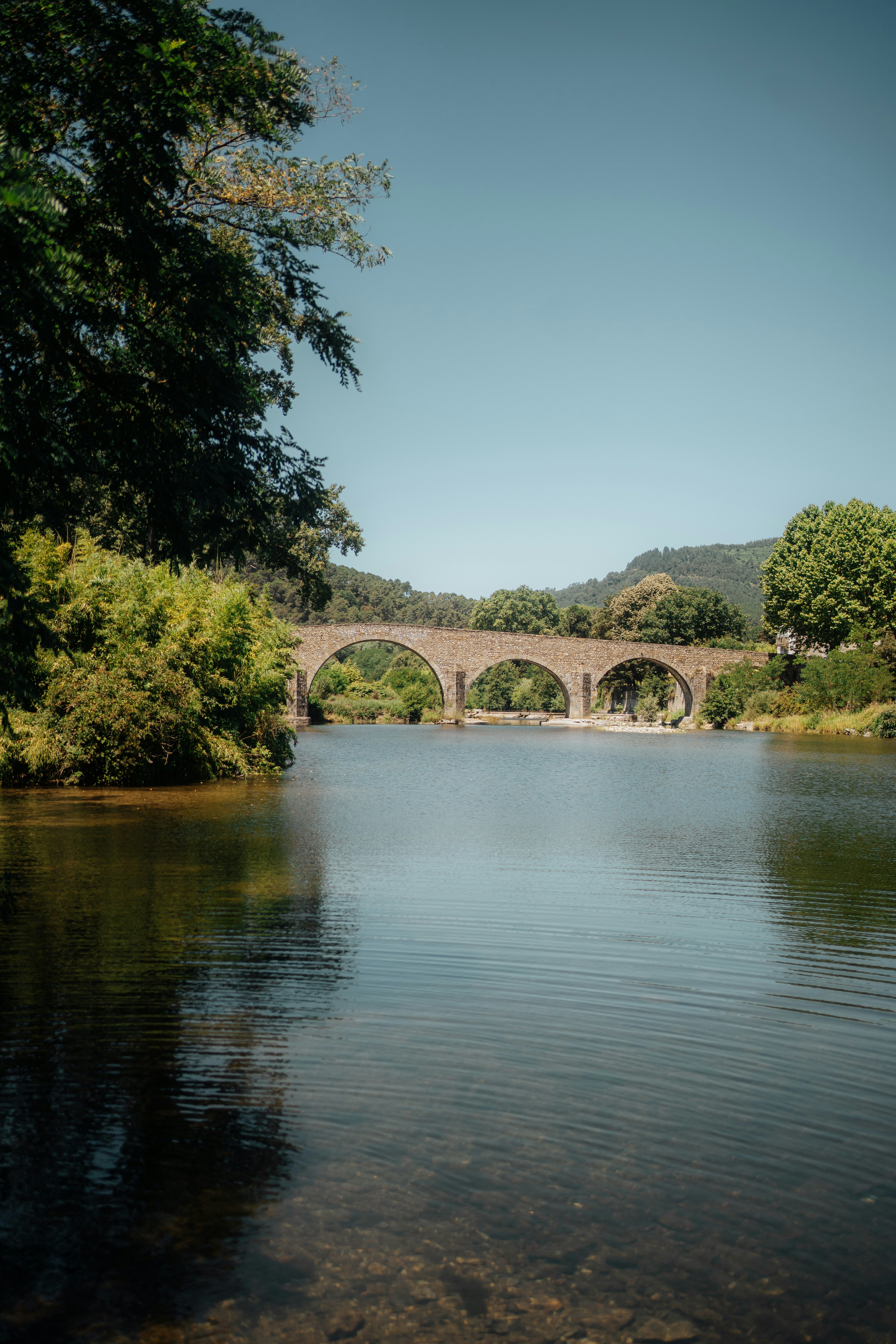 Stone bridge arches gracefully over a serene river, surrounded by lush greenery and distant hills.