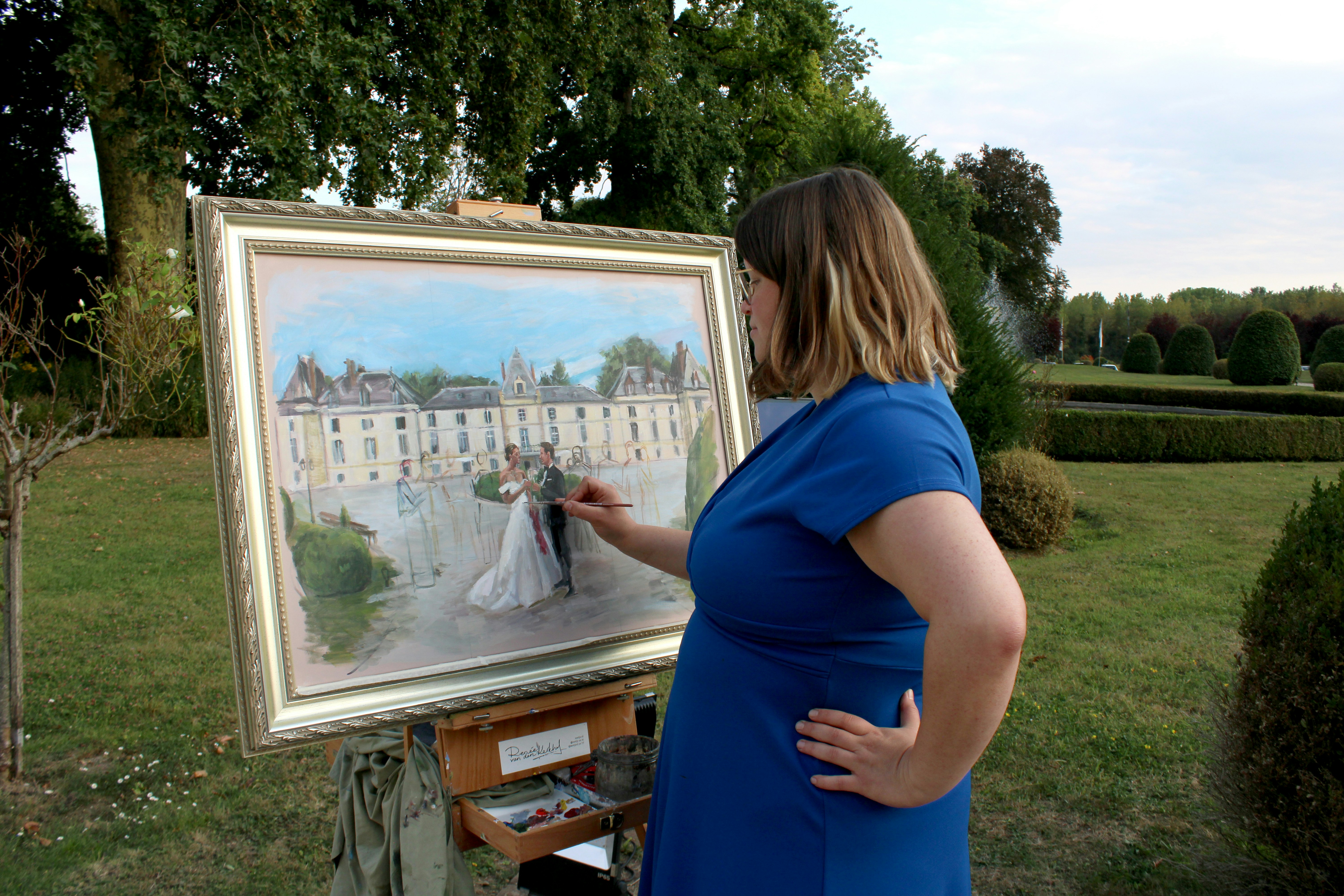 Destination wedding in France: live painter Renée wearing a blue maxi dress, painting an outdoor wedding scene on location with the couple in the center. The setting of the artwork is in front of Château D'aveny, a luxury 18th century castle. The artist is working with a paintbrush and a foldable French box easel. Castle gardens behind the easel. | Woman painting a wedding scene outdoors at a chateau.
