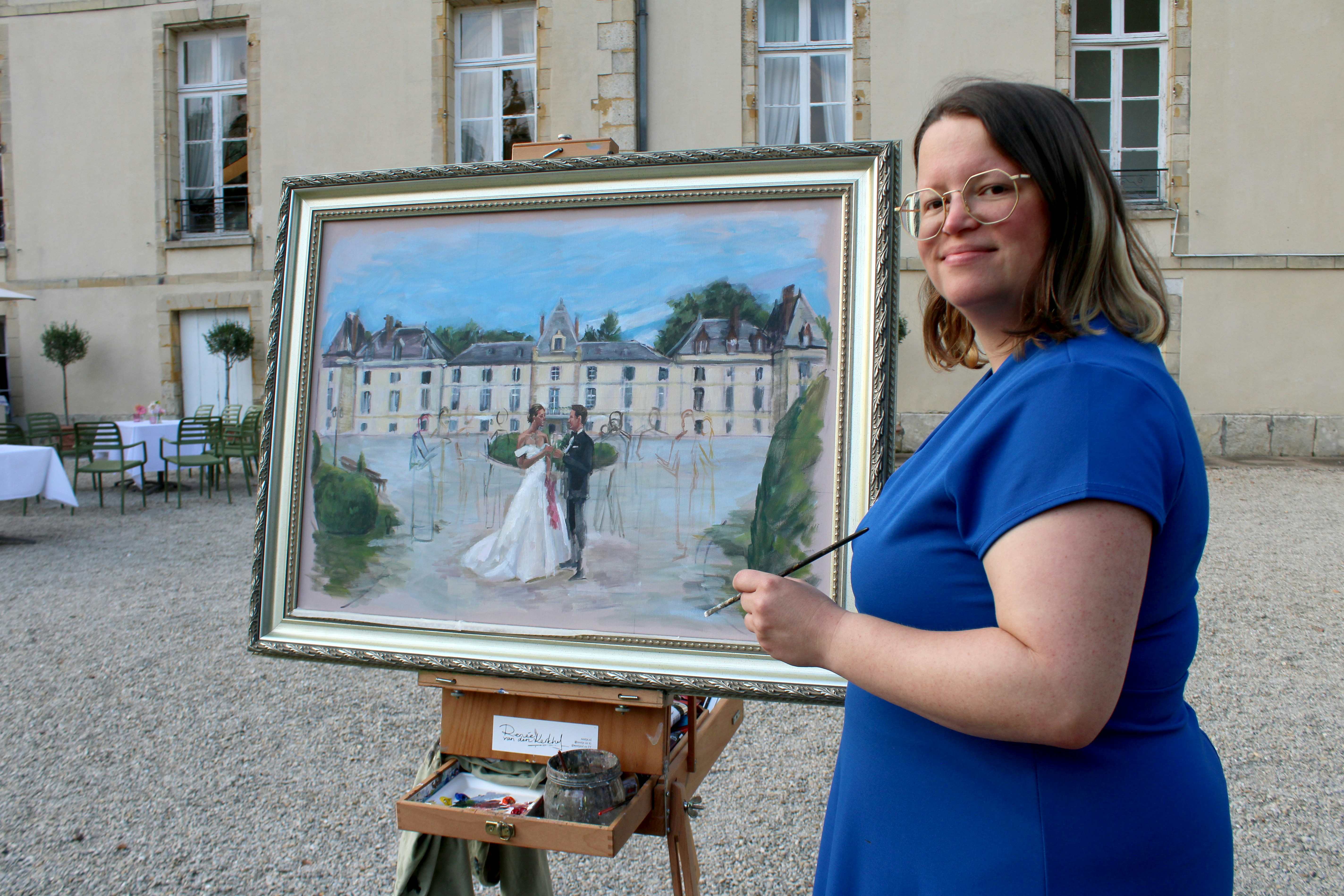 Live wedding painting in a decorative frame, work in progress, The background and the wedding couple have a few details but the other people are still rough sketches. Live painter Renée looks into the camera, wears a blue maxi dress, painting an outdoor wedding scene with the couple in the center. The setting of the artwork is in front of Château D'aveny, a luxury 18th century castle. The artist is working with a paintbrush and a foldable French box easel. In the background we see part of the castle.