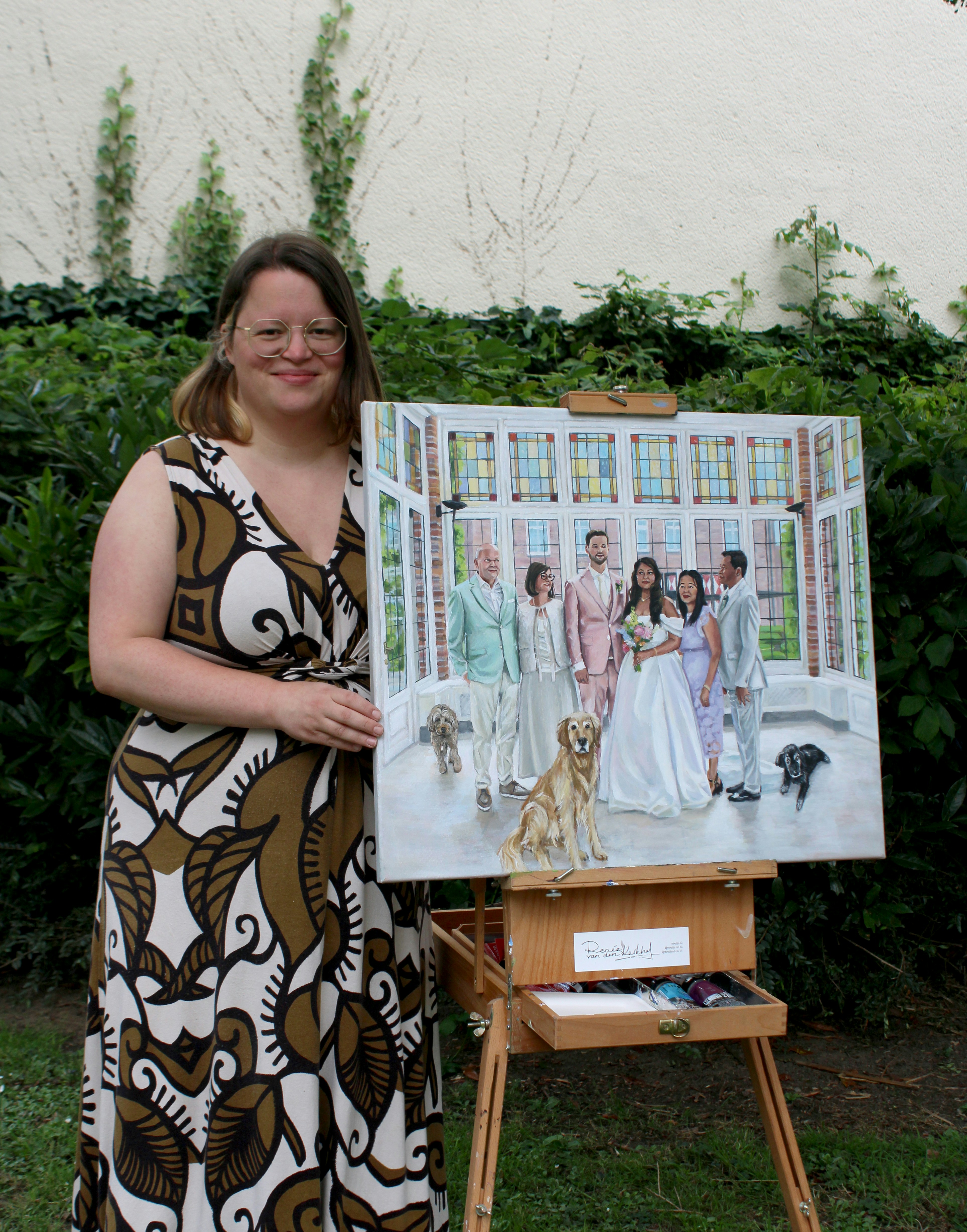 Live wedding painter Renée showing a finished wedding painting, that started live on location. Family portrait with parents and dogs. The groom is wearing a pink suit, the bride a big off-shoulder white dress with bows. The setting is indoors, in the greenhouse of Maurick Castle. The painting rests on an easel and the female artist stands next to it, in a dress with white, brown and black patterns. | Woman holds a wedding portrait painting with family and dogs