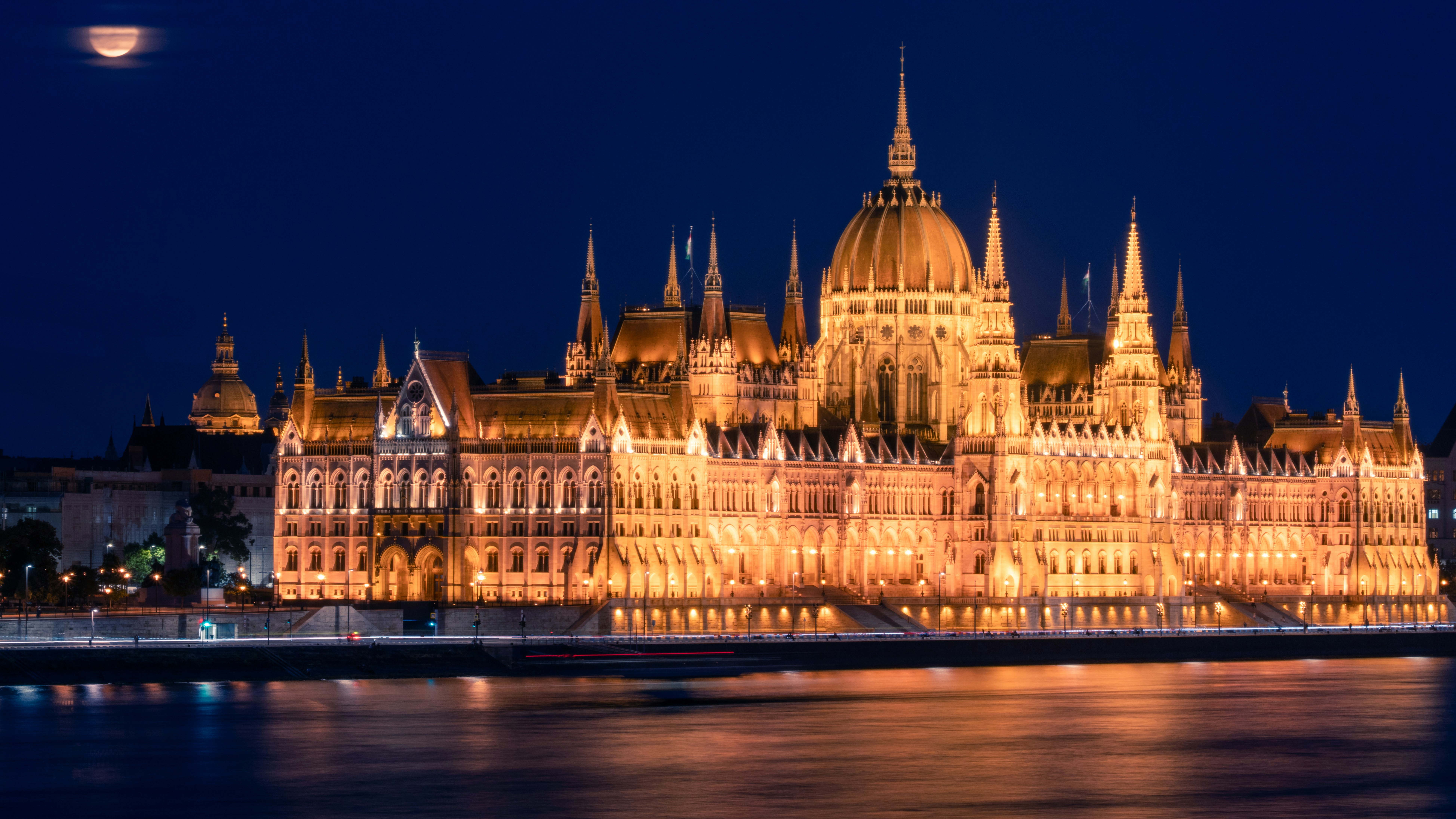 The Hungarian Parliament Building in the night with the moon half-covered with cloud. | Ornate building illuminated at night with moon