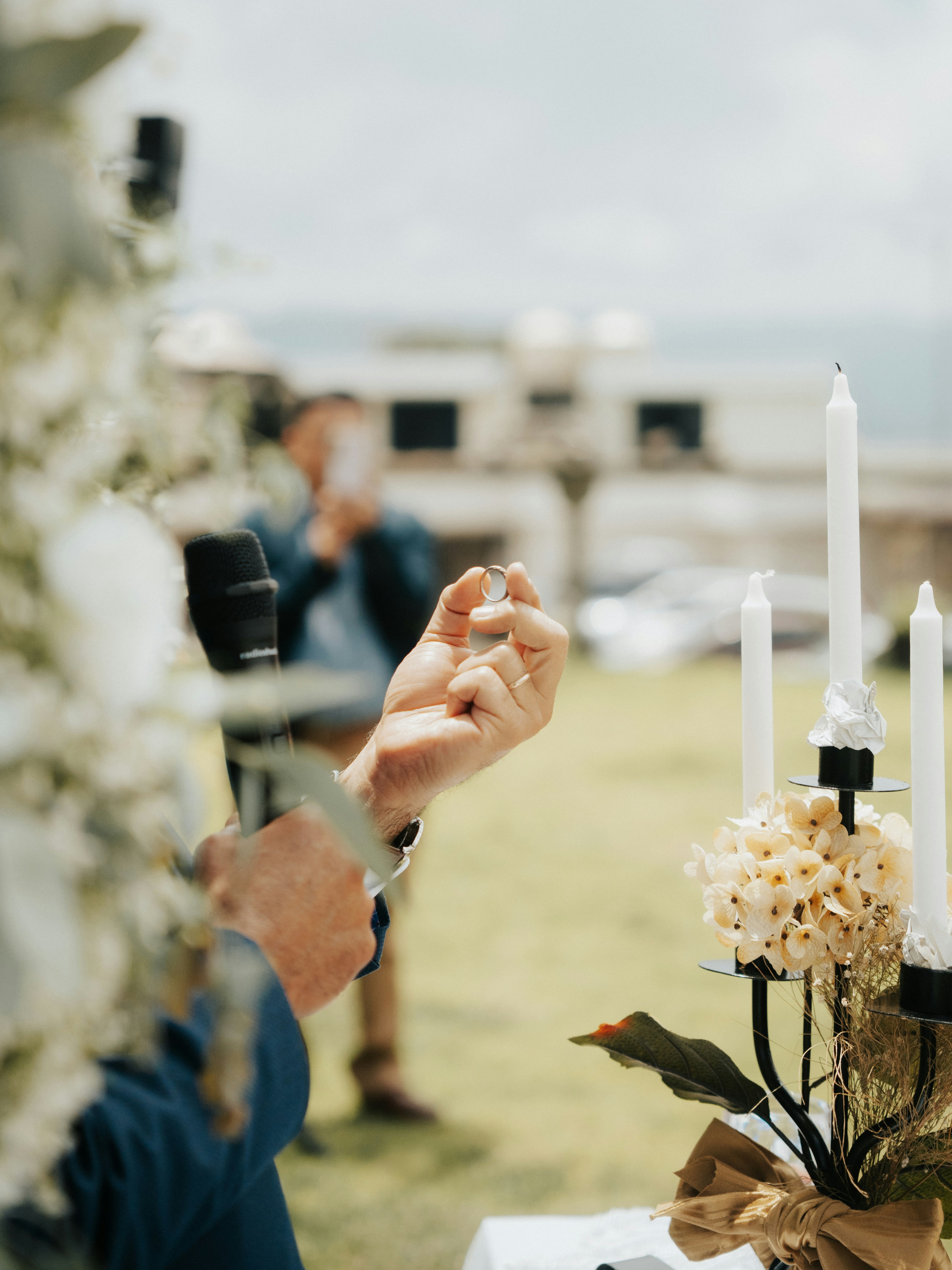 A hand delicately holds a wedding ring during a ceremony, surrounded by floral arrangements and candles. The scene captures the emotional significance of the moment.