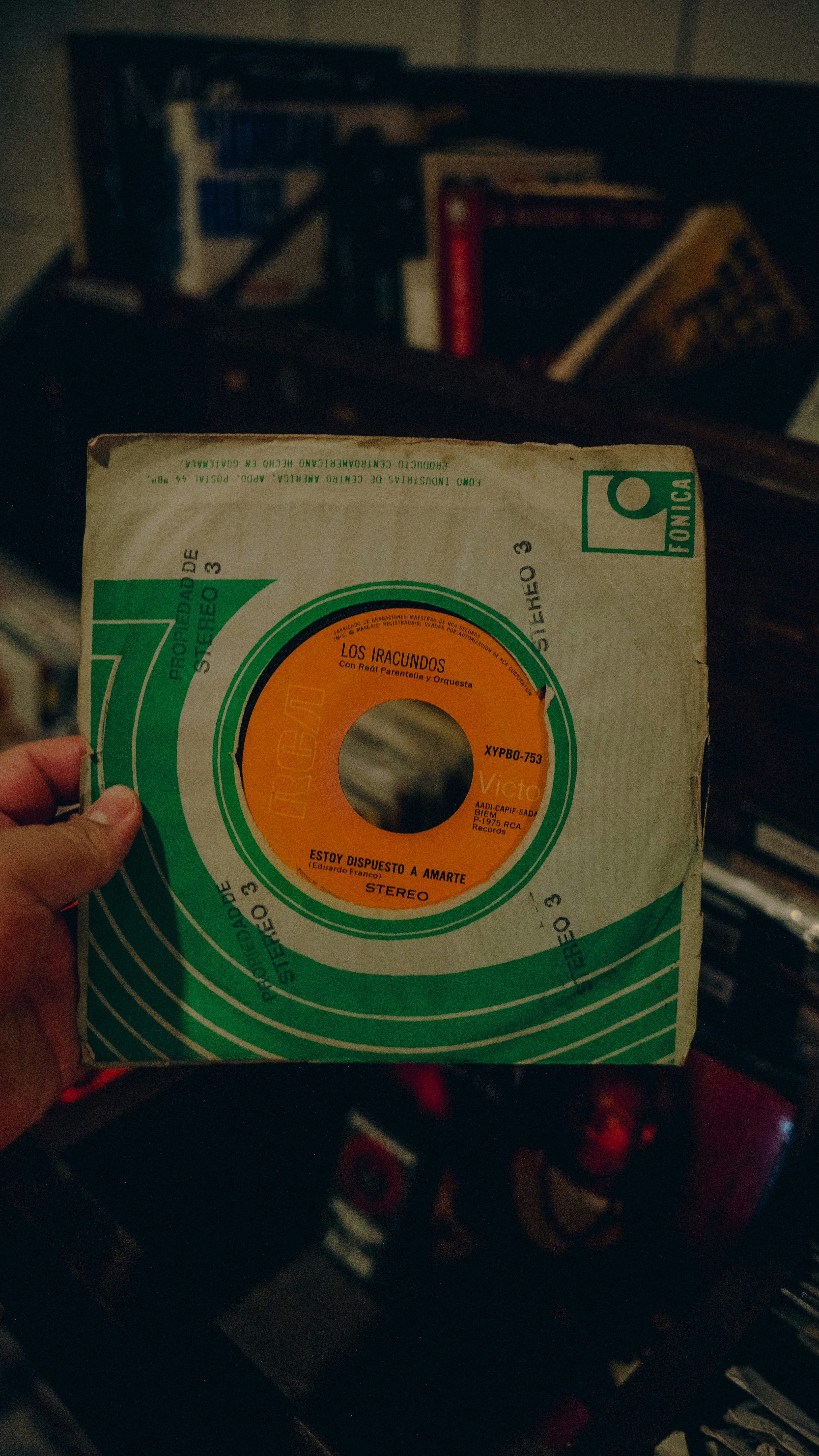 A hand holding a vintage record sleeve featuring the band Los Iracundos, surrounded by a backdrop of books and other records. The design reflects a retro aesthetic.