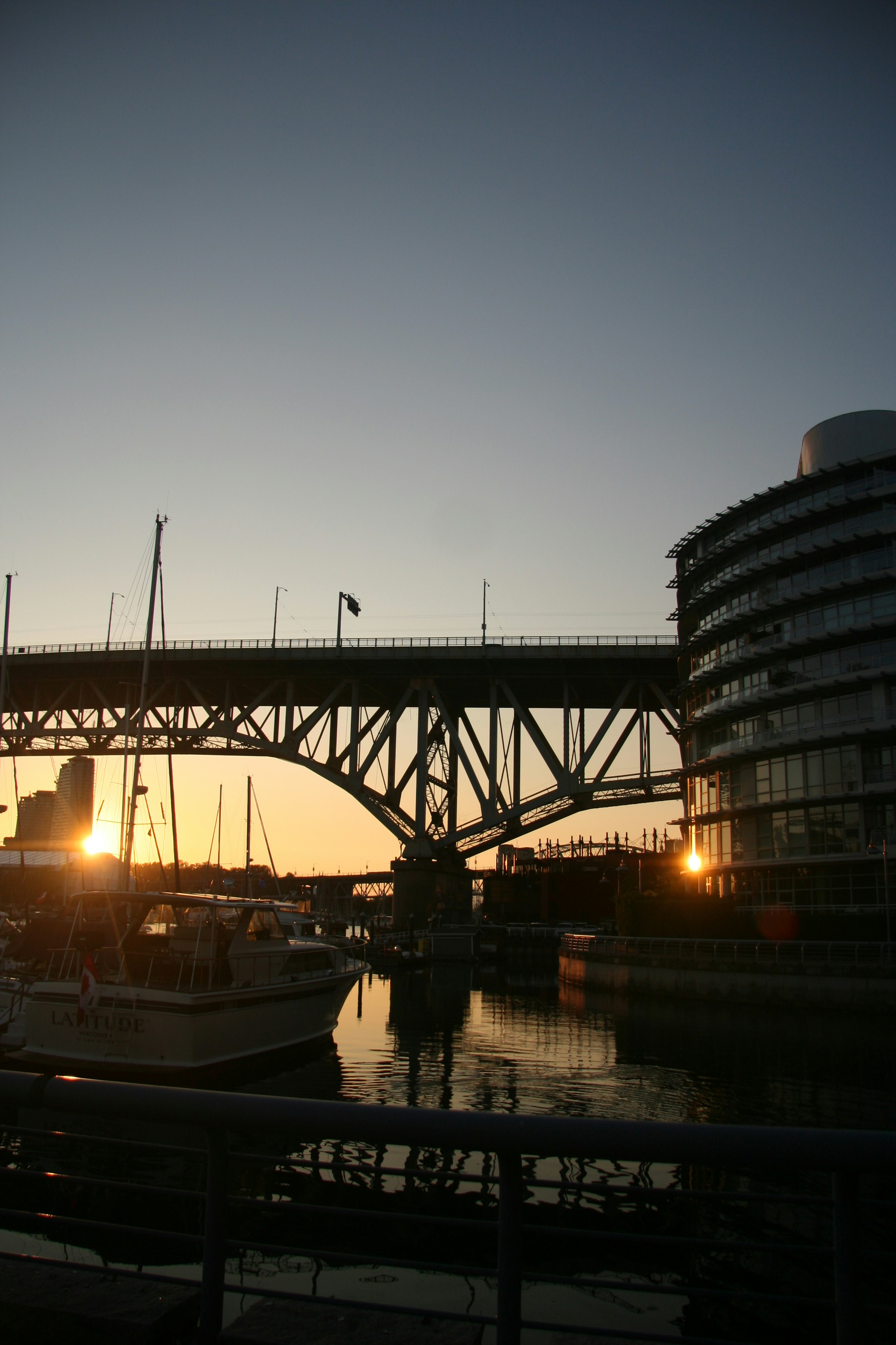 Silhouette of a bridge against a vibrant sunset, with reflections shimmering on the water's surface and boats gently bobbing in the harbor.