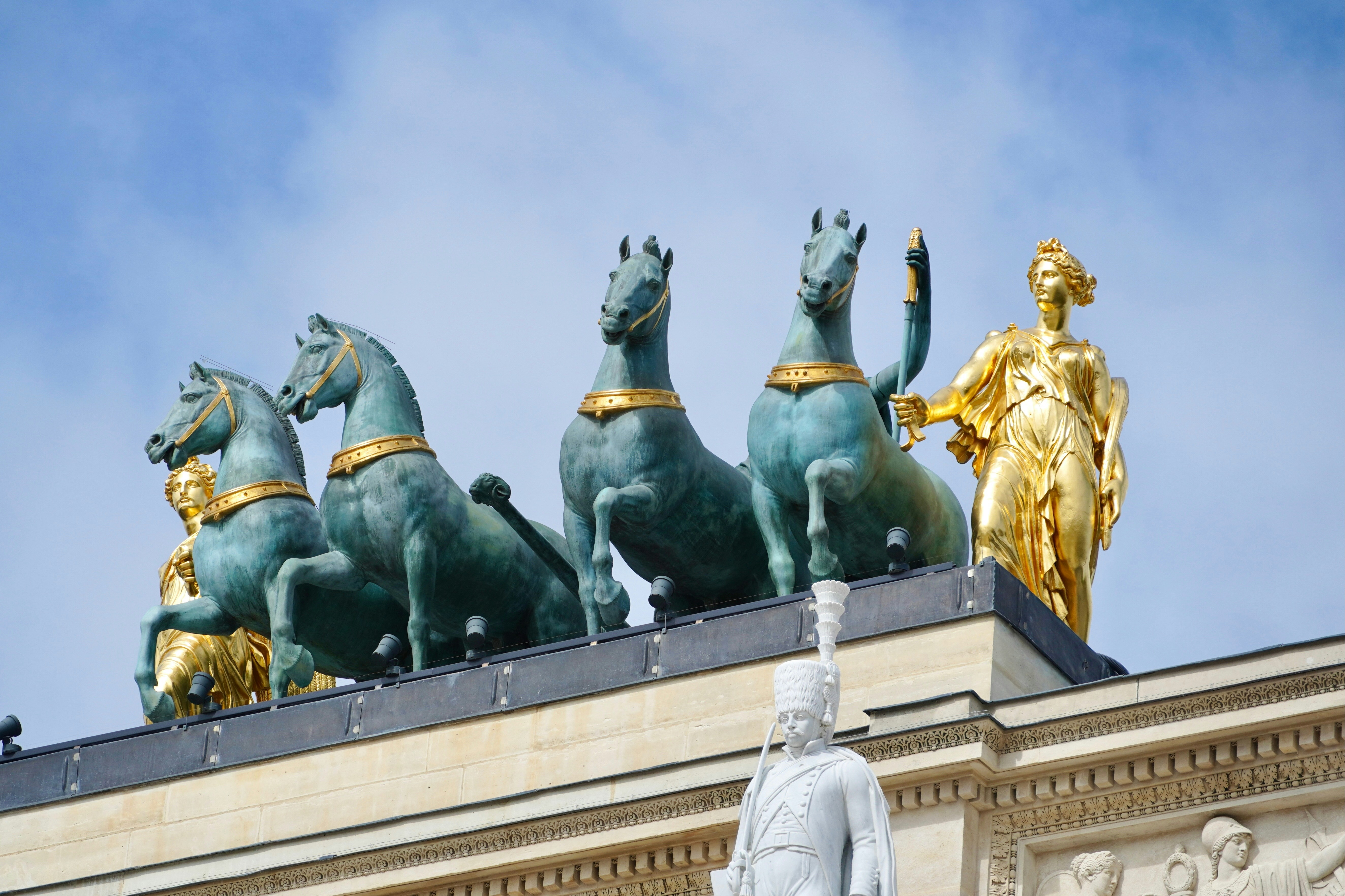 Bronze horses and a golden figure atop a grand architectural structure against a blue sky.