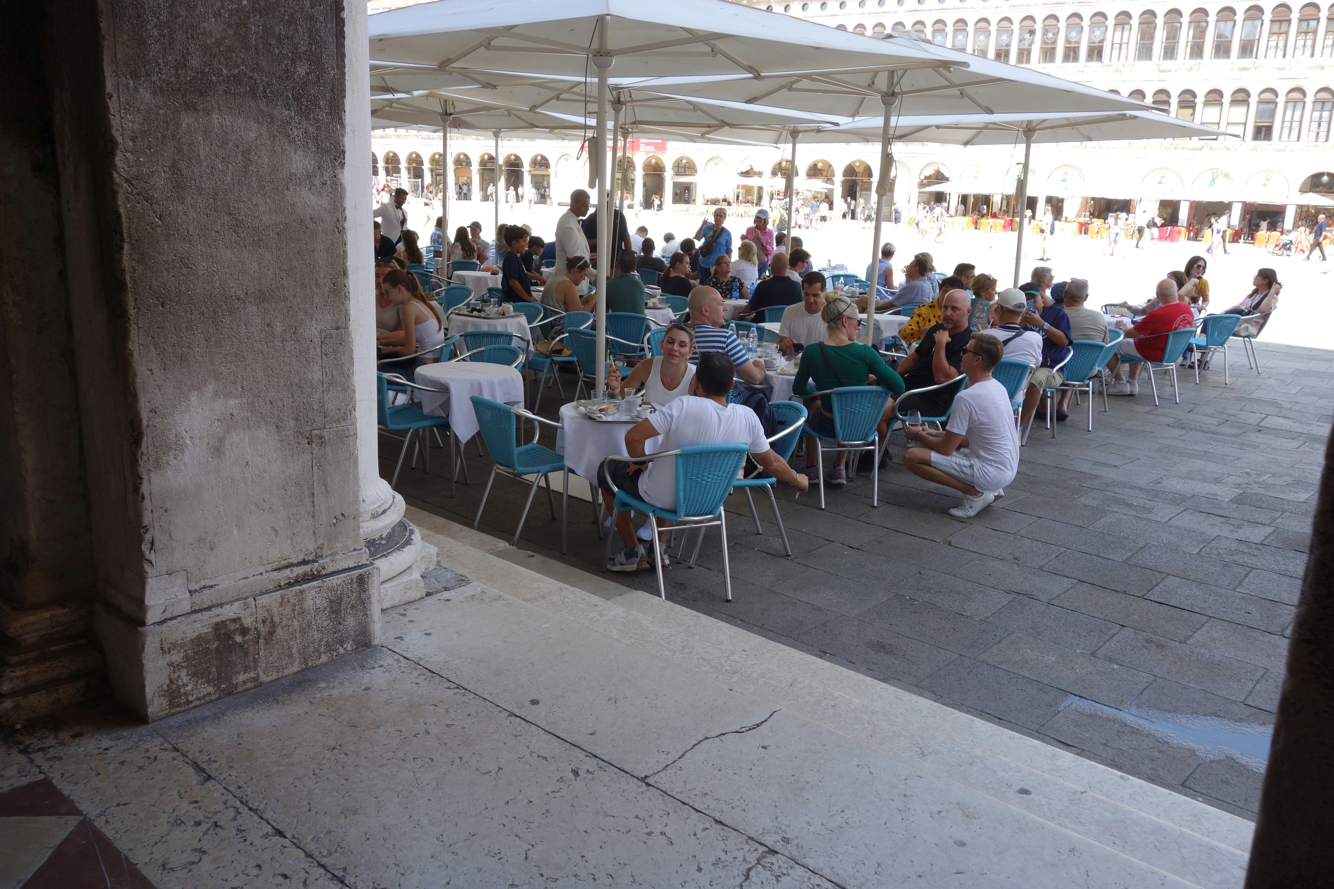 Crowd enjoying refreshments at outdoor café in a bustling Venetian square, with a historic backdrop. 