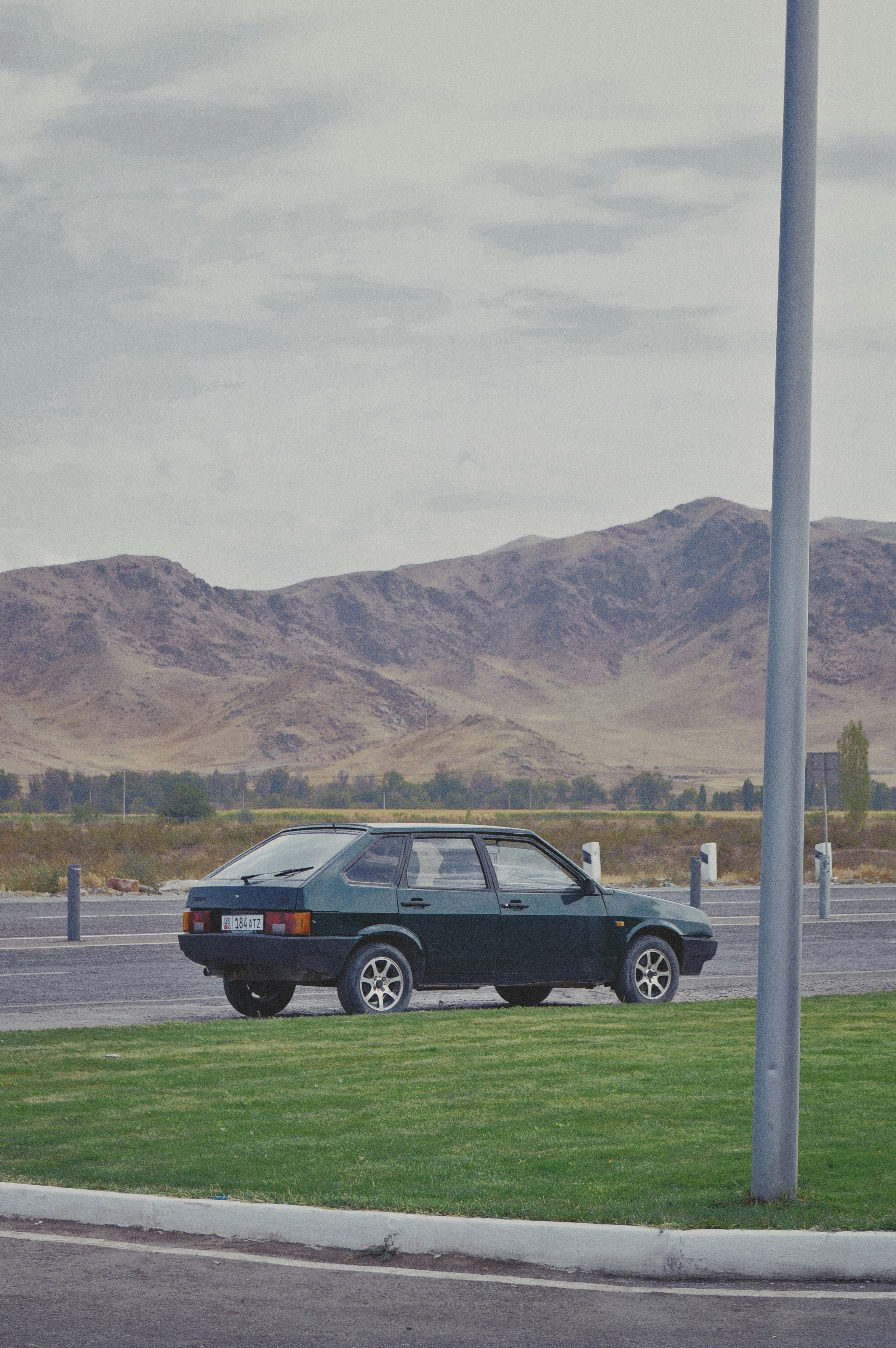 A blue-green sedan sits parked in a sparse desert parking lot with mountains rising on the horizon. A tall streetlamp foregrounds the scene, framing the quiet composition.