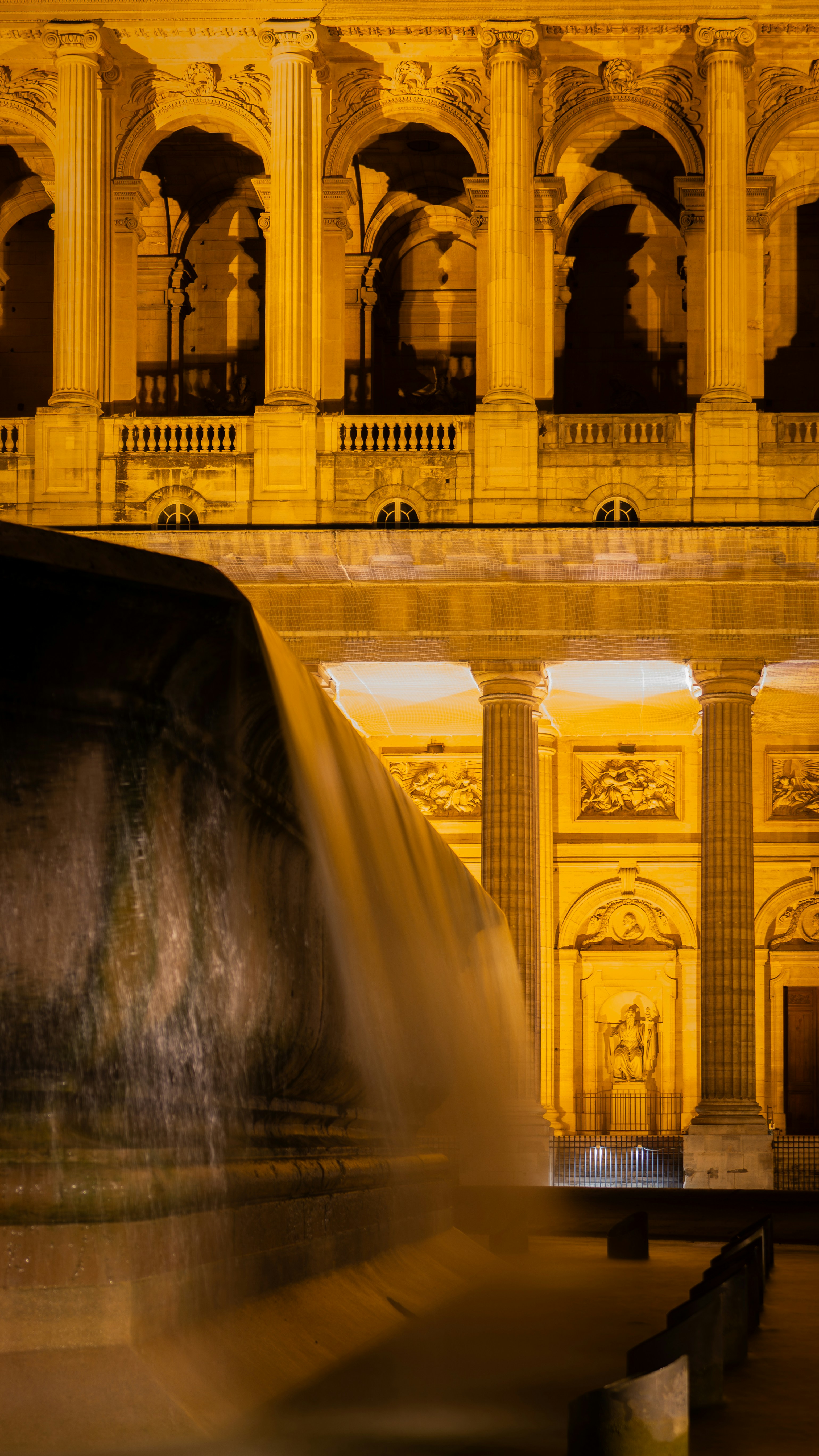 Illuminated fountain cascading in front of ornate classical architecture at night.
