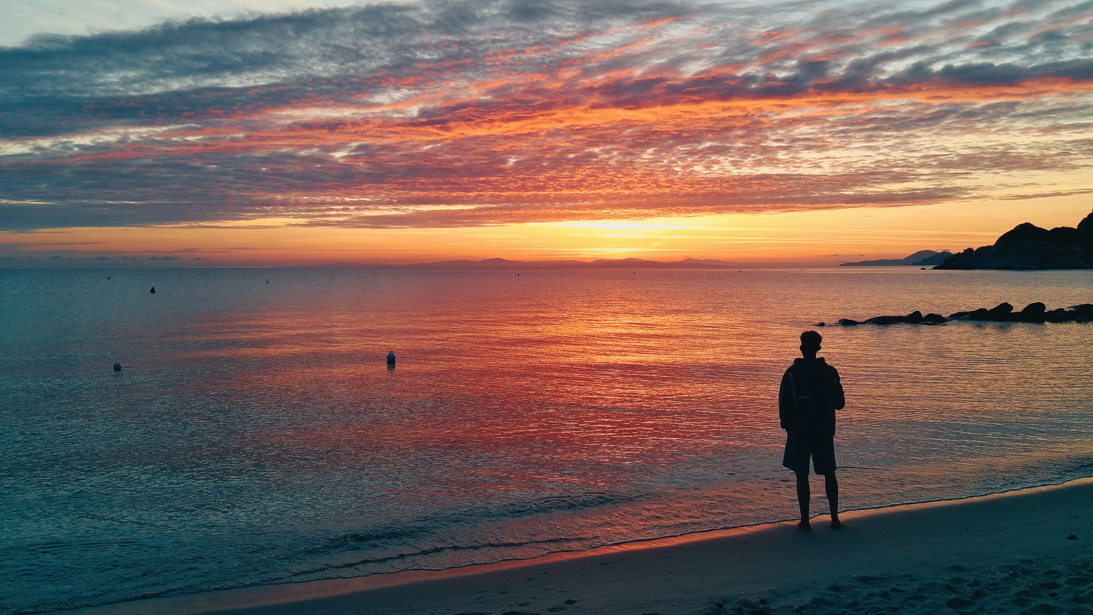 Silhouette of a person standing on a beach, gazing at a vibrant sunset reflecting on calm waters.