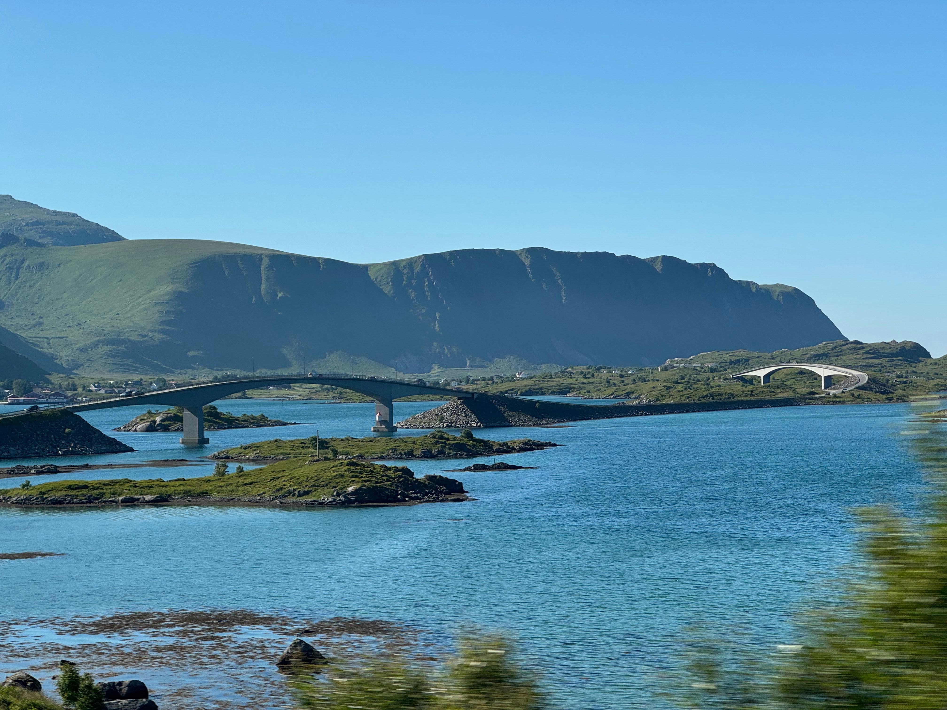 Two elegant bridges arching over a serene body of water, framed by lush green hills under a clear blue sky.