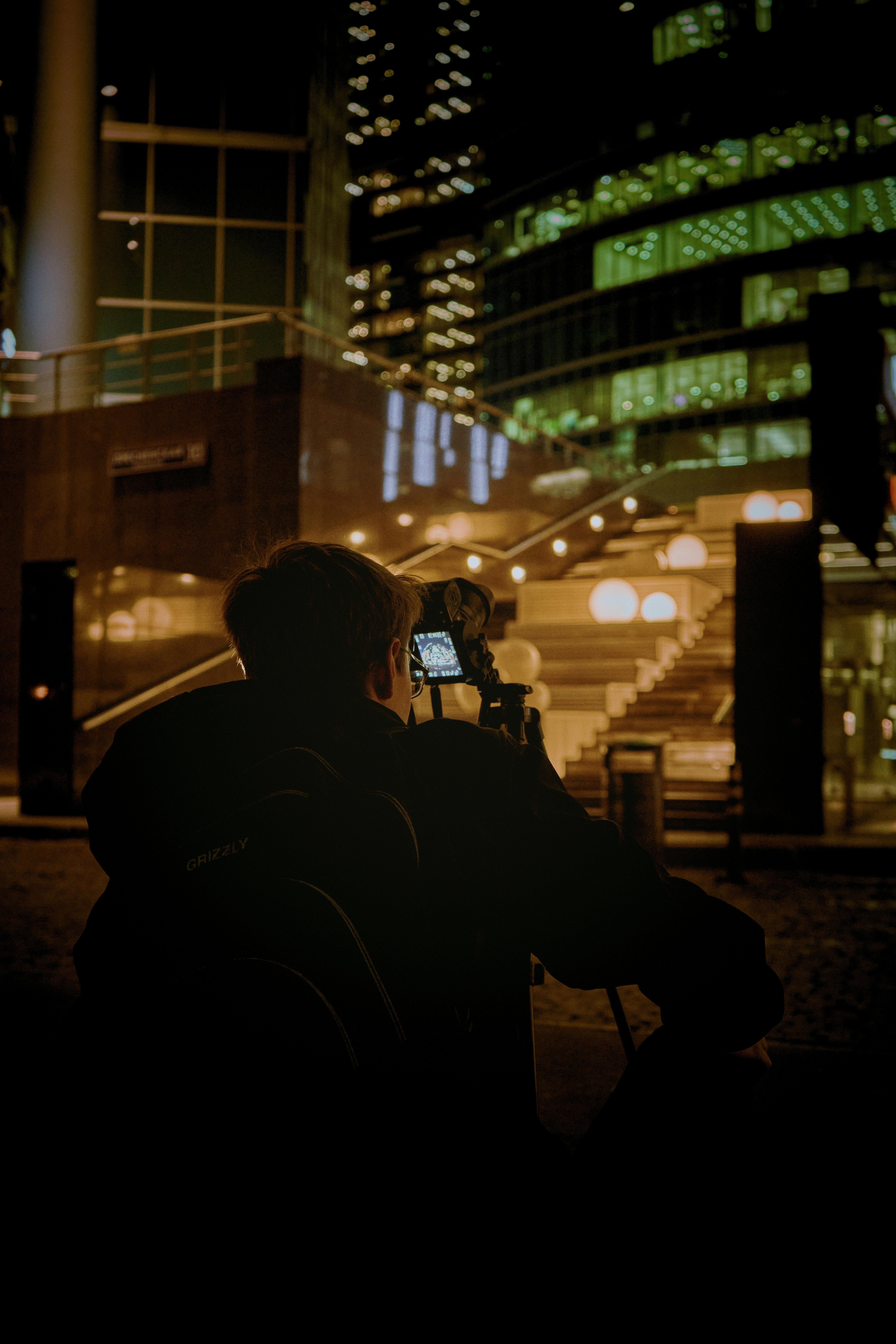 A photographer, silhouetted against a modern building, focuses on capturing the vibrant nighttime scene with their camera. The illuminated architecture creates a dynamic backdrop.