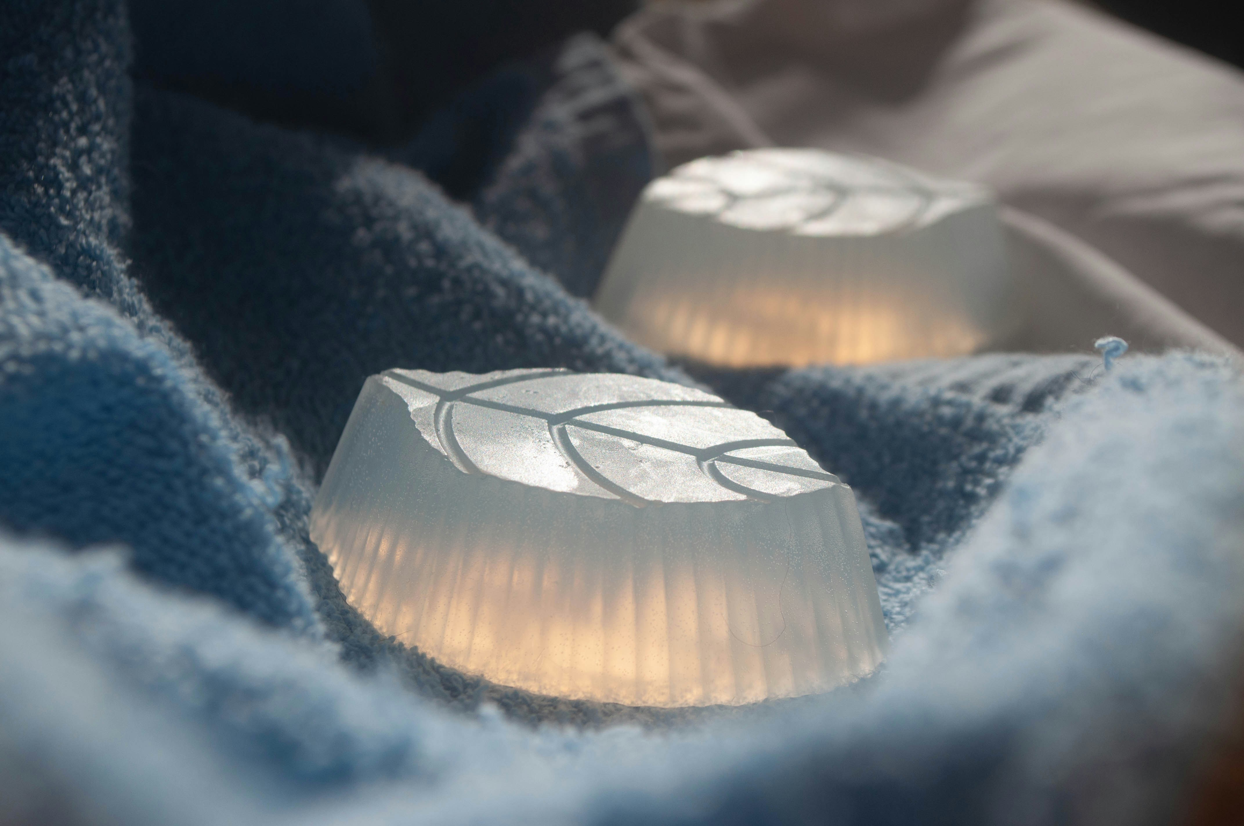 Two transparent leaf-shaped glycerin soap bars on a blue towel background | Two translucent soaps with leaf imprints resting on towels