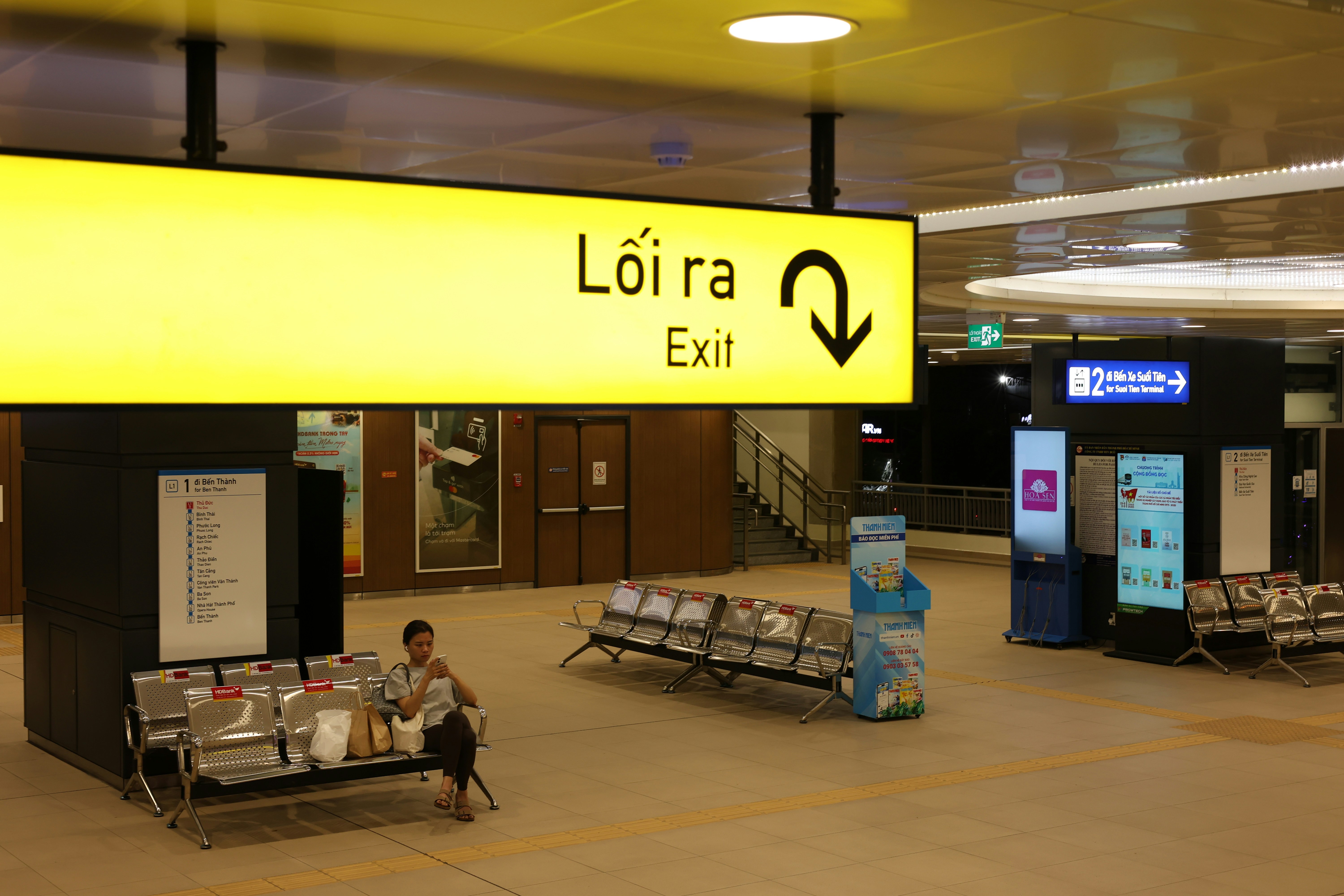 Bright yellow exit sign illuminates a quiet waiting area in a transit station, with a lone figure seated on metal benches.