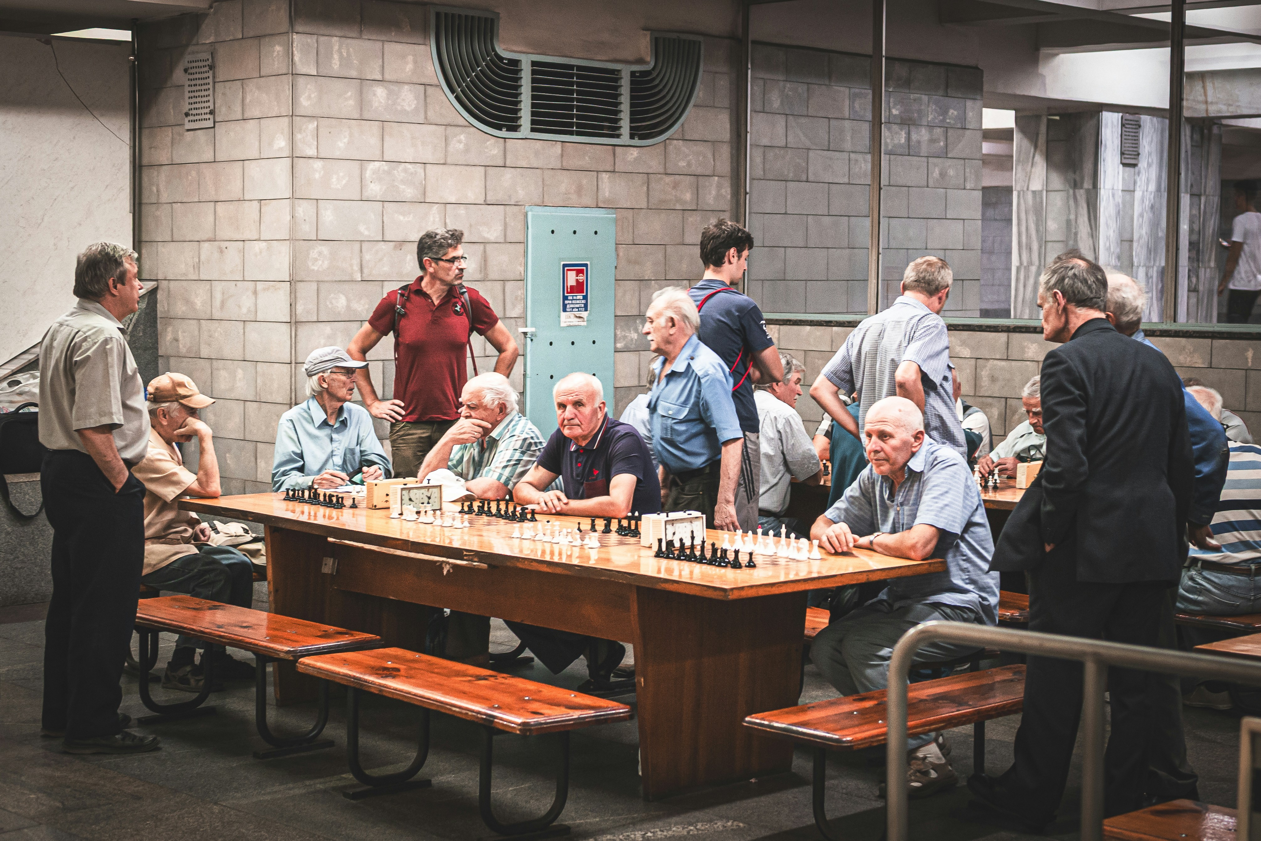 A group of elderly men playing chess at a long table in a public indoor space. A candid documentary moment of community and everyday life. | Men playing chess at tables in a room