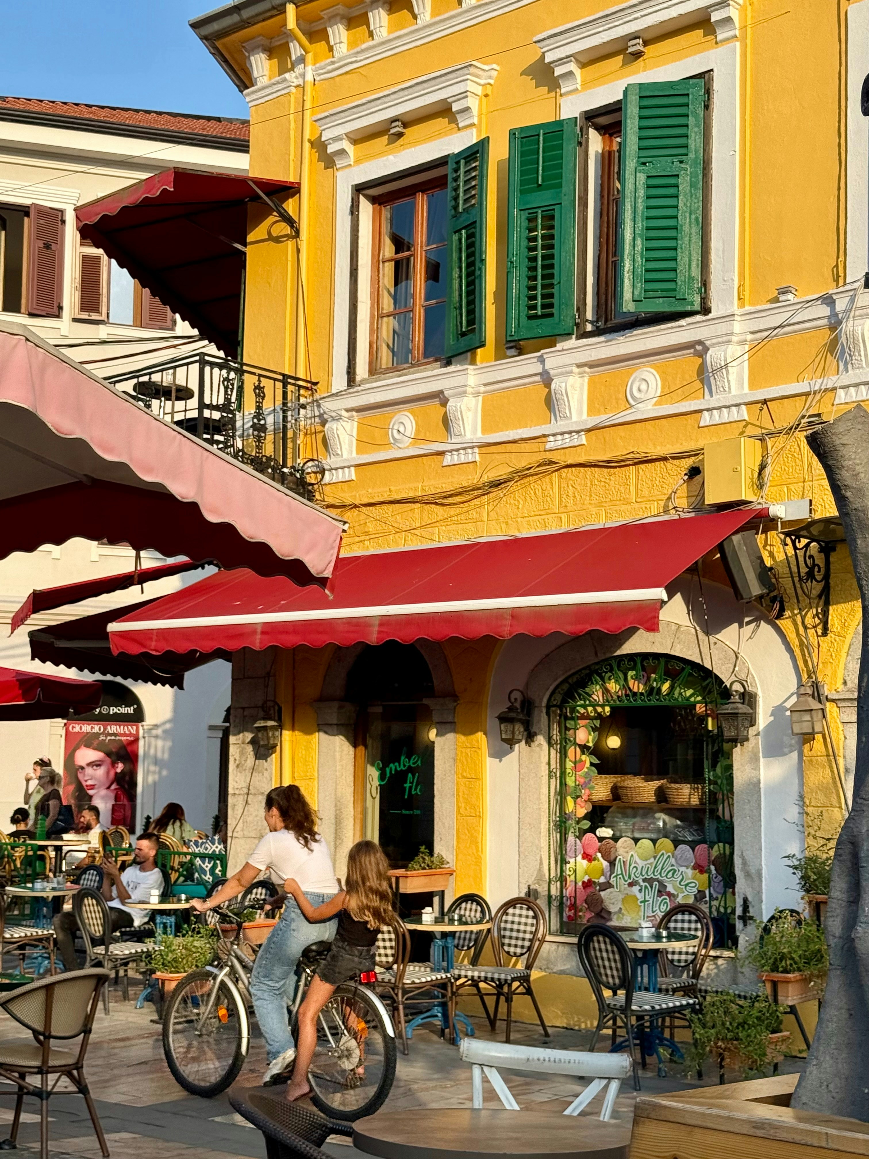 Colorful | Two women ride bicycle past outdoor cafe
